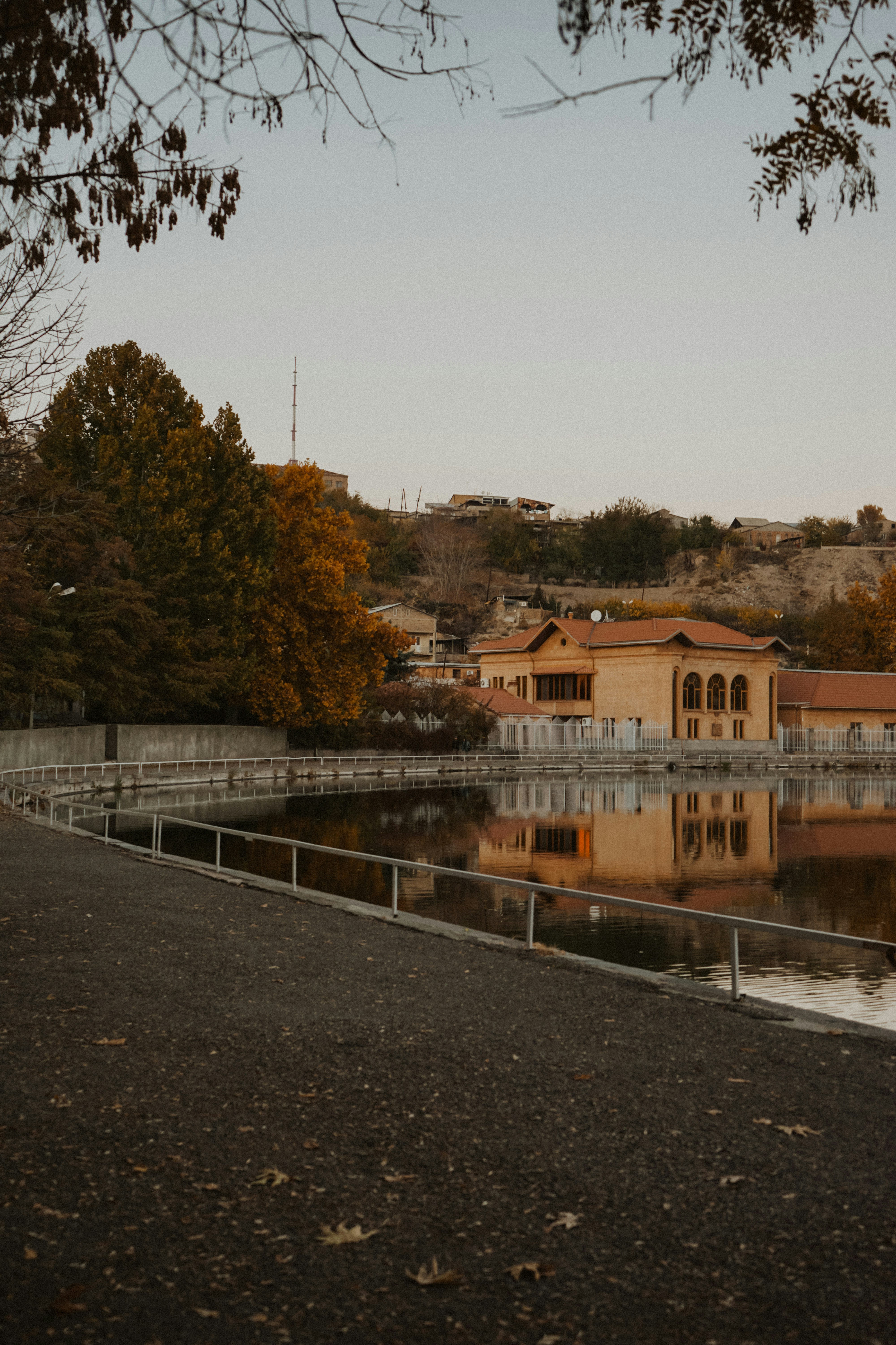 Lakeside view with autumn foliage and reflections of buildings on calm water under a soft sky.