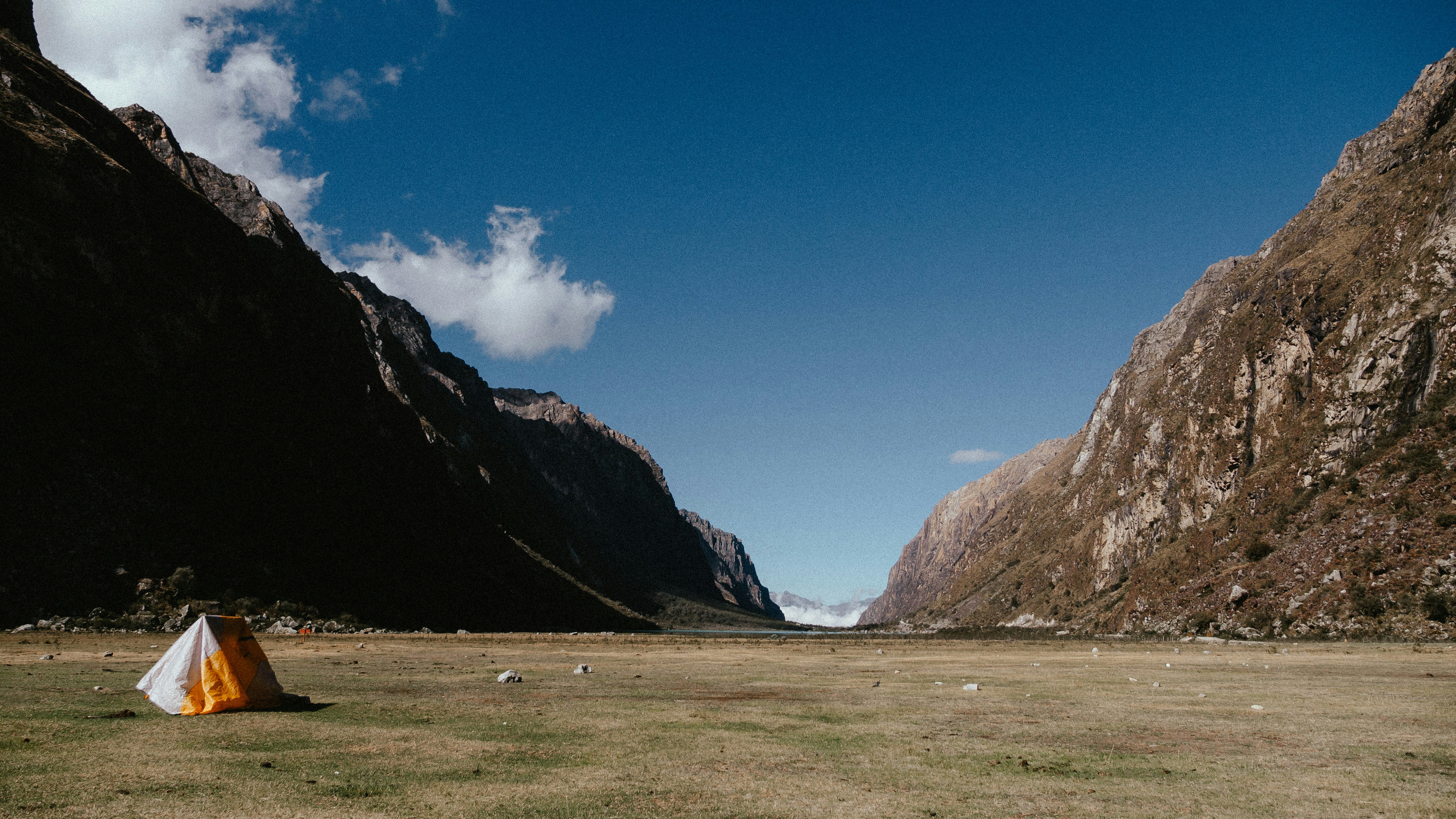 Tent pitched on a vast grassy plain surrounded by towering mountains under a clear blue sky.