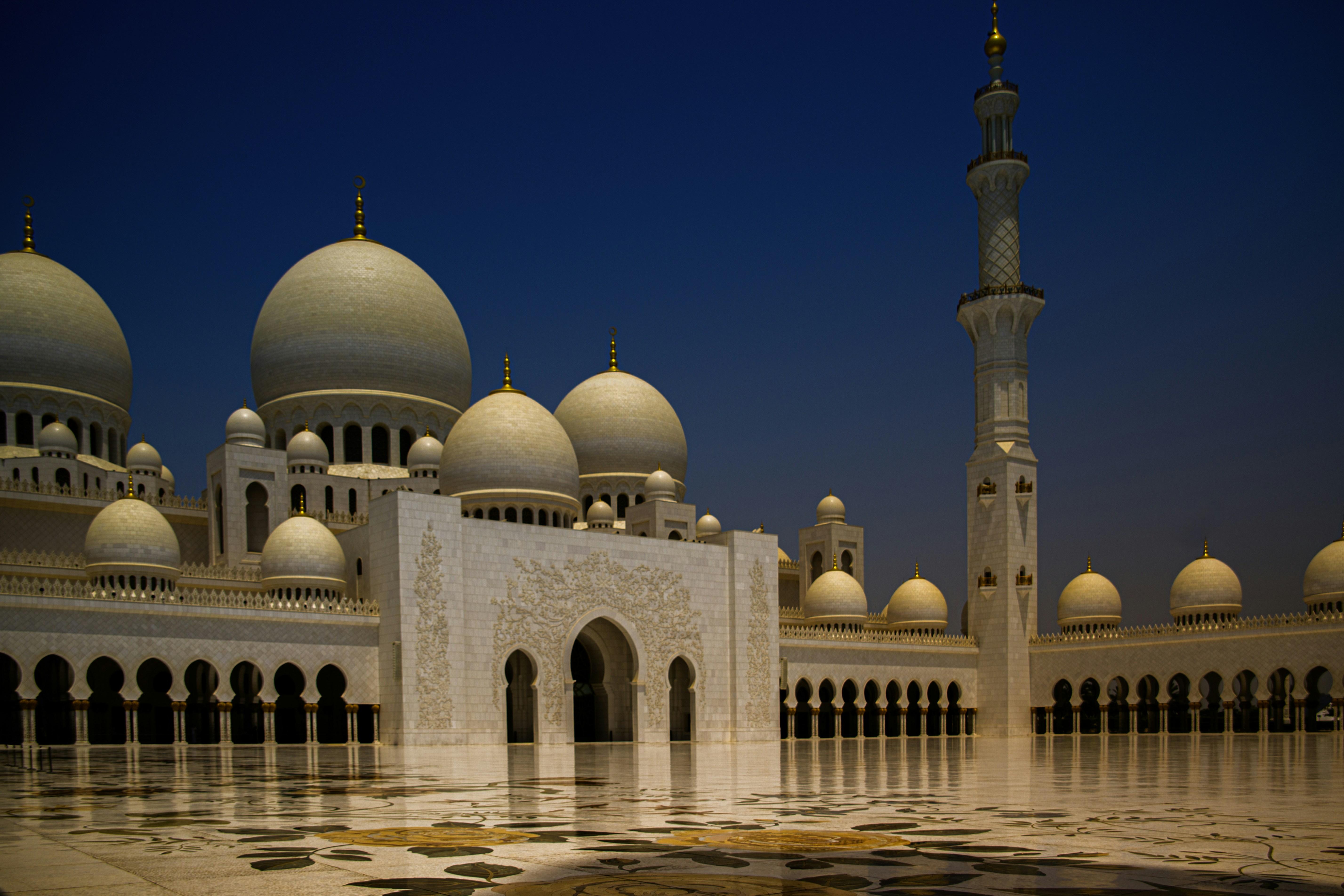 Illuminated domes and minaret of Sheikh Zayed Mosque reflecting on polished courtyard under a deep blue evening sky.