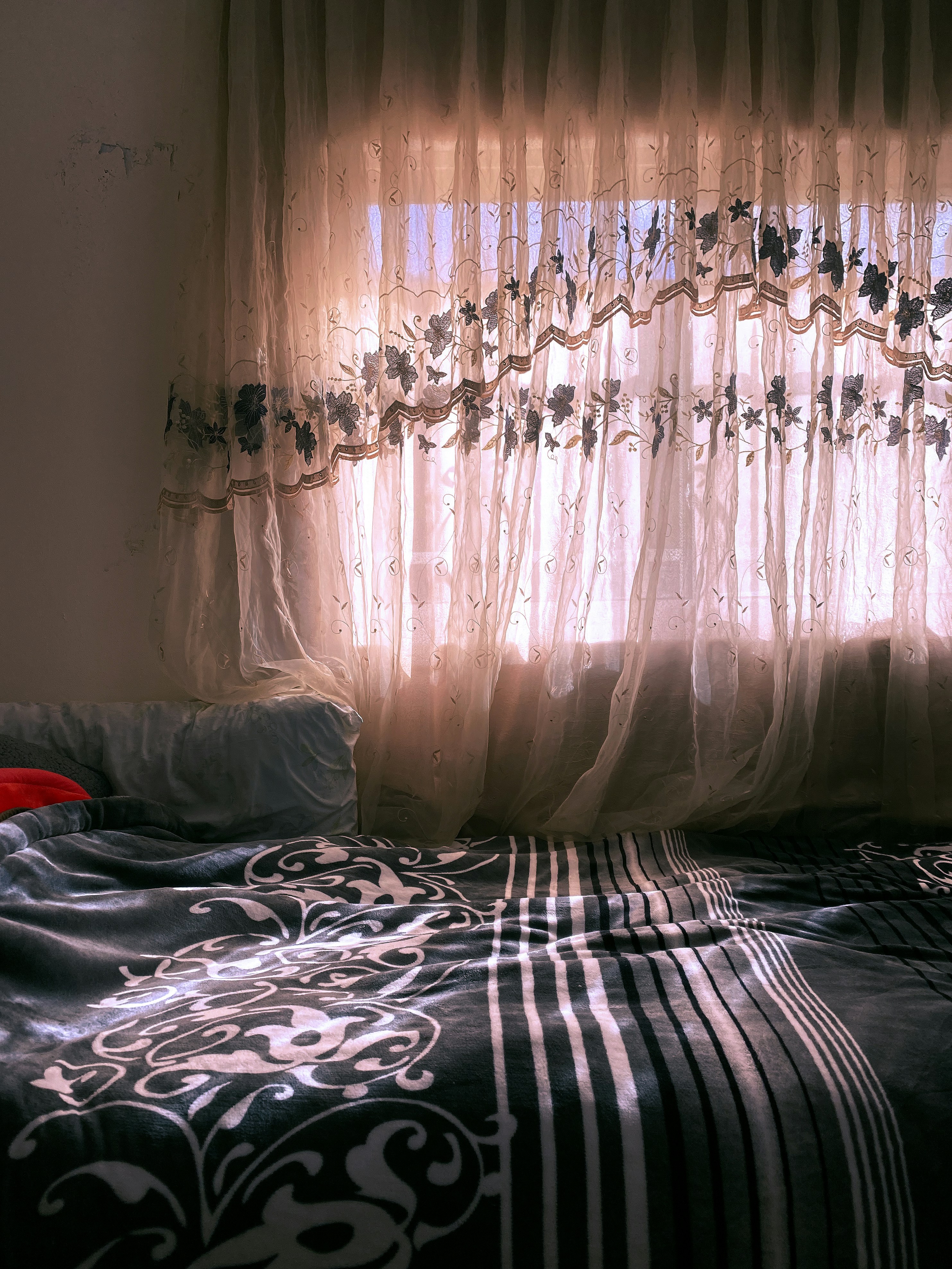 Sunlight glows through sheer embroidered curtains, casting a warm, hazy glow over a neatly made bed with a bold patterned coverlet.