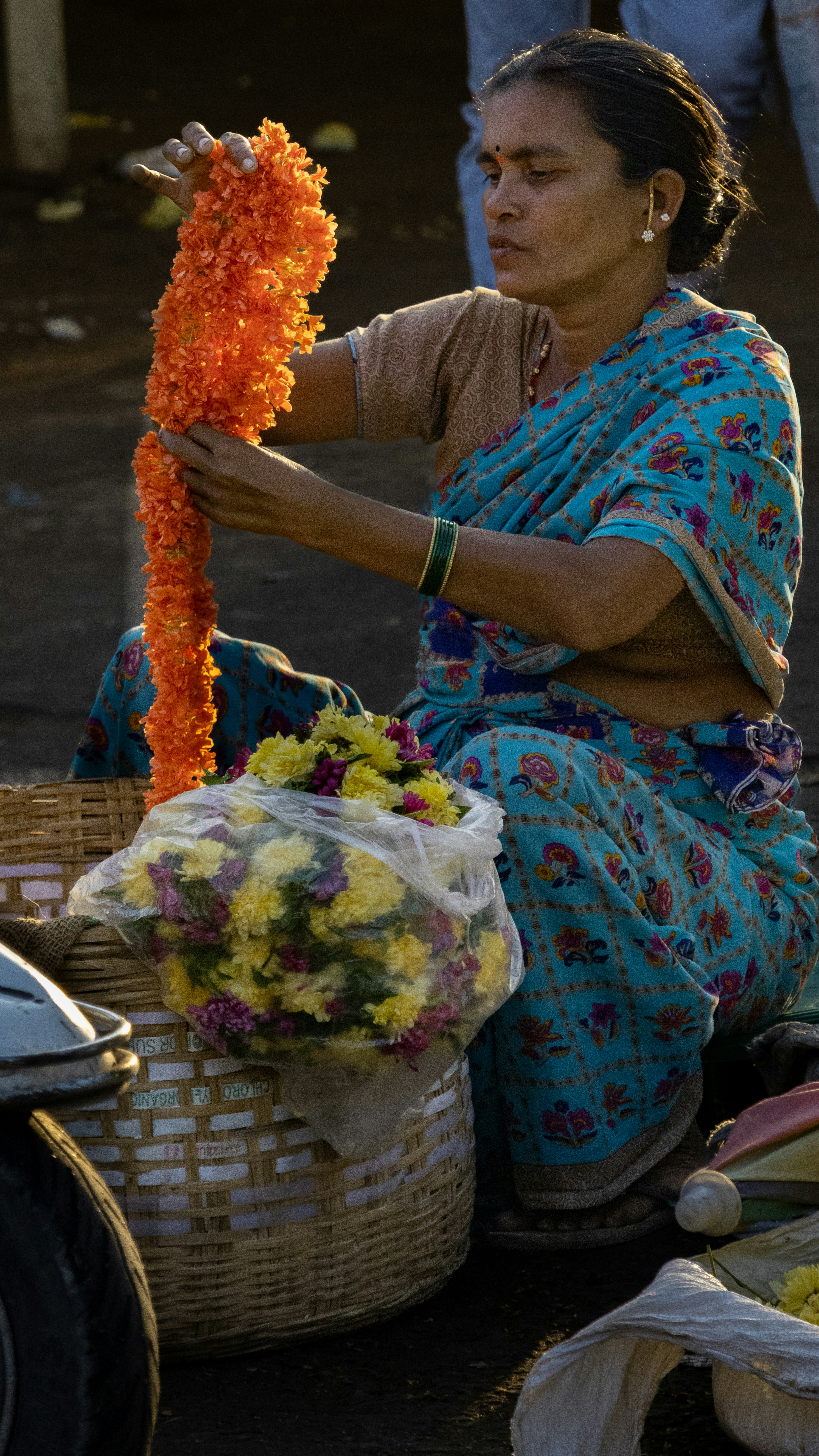 Sunlit Garland Craft: A Woman Weaving Orange Flowers at a Street Market