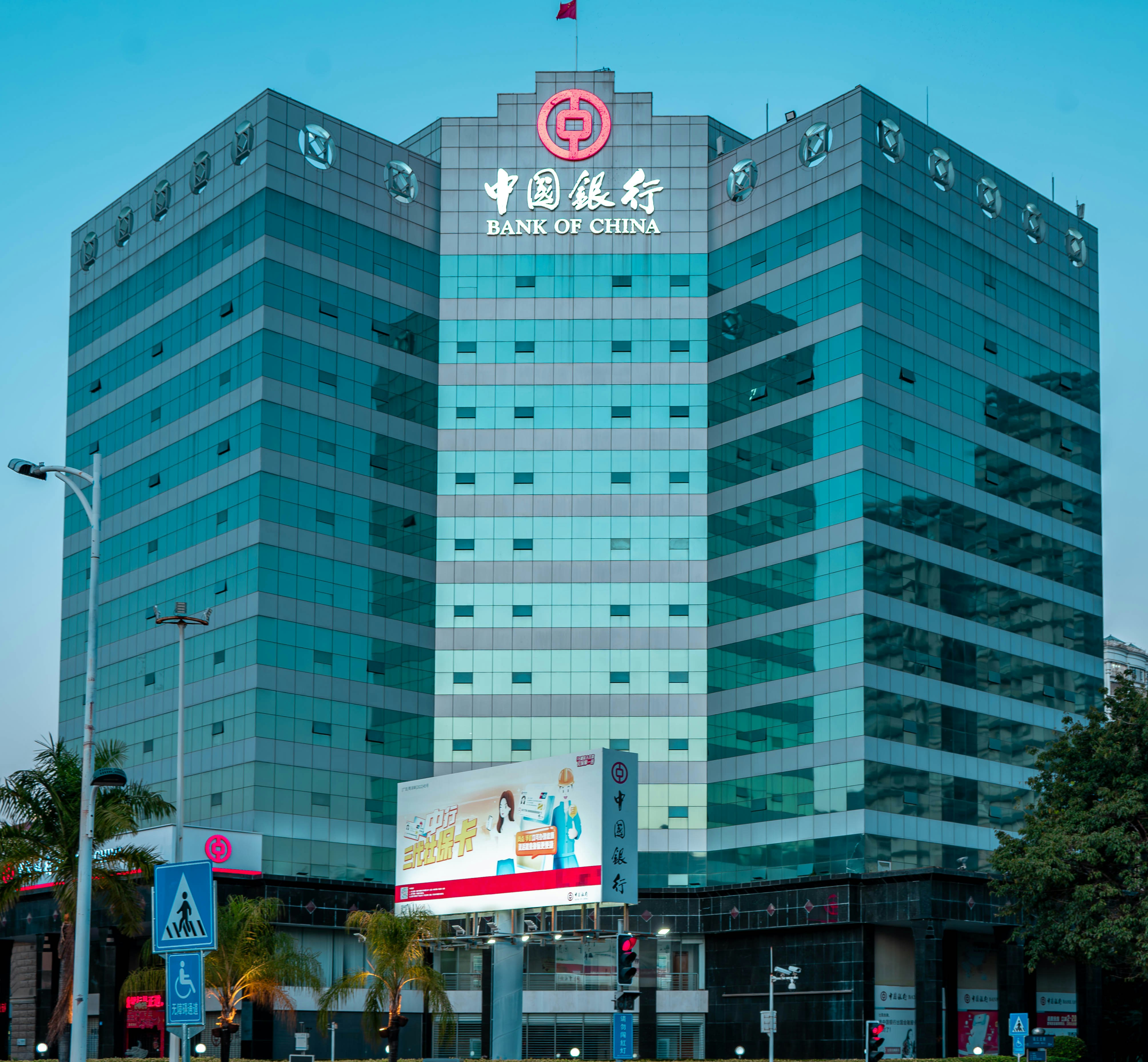 The modern glass facade of the Bank of China building glows in the cool light of dusk. Its prominent illuminated logo and Chinese characters (中国银行) stand out atop the sleek, multi-story structure. Situated in an urban environment, street signs and palm trees frame the base of the impressive corporate building.