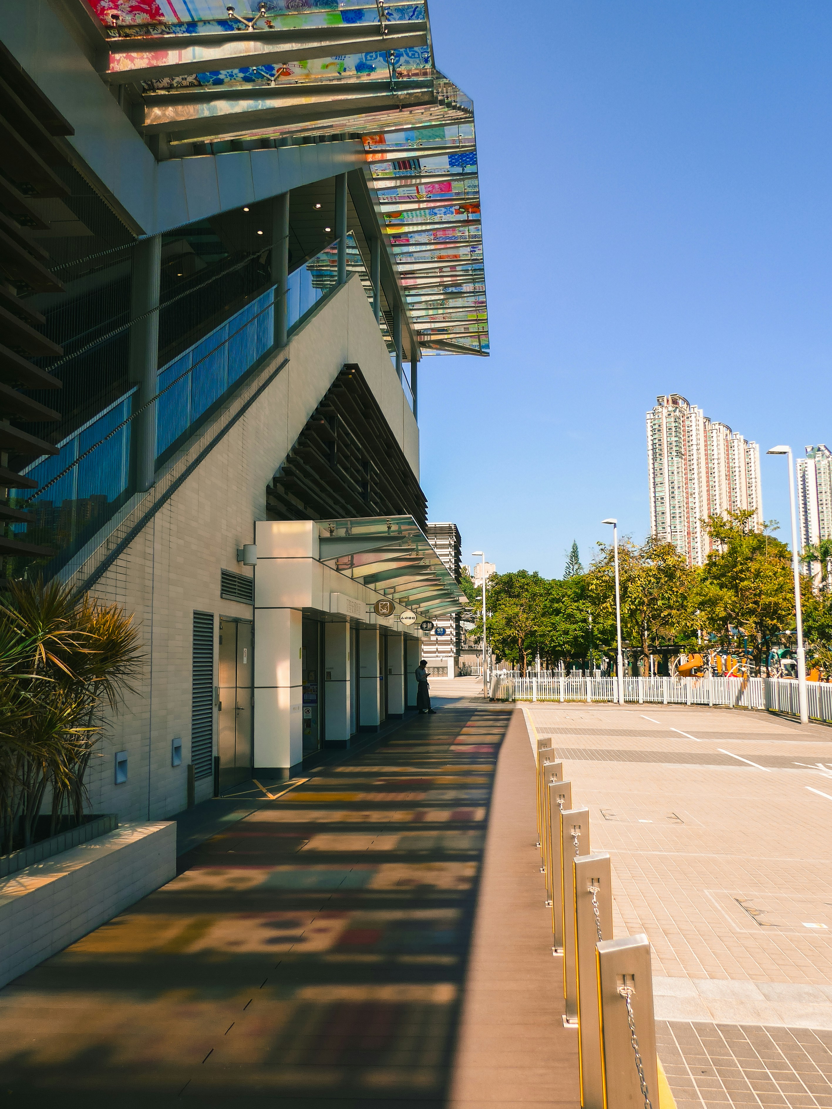 Sunlit, glass-fronted building lines a long walkway with bold shadows. Distant skyline rises beyond trees, framed by modern architecture.