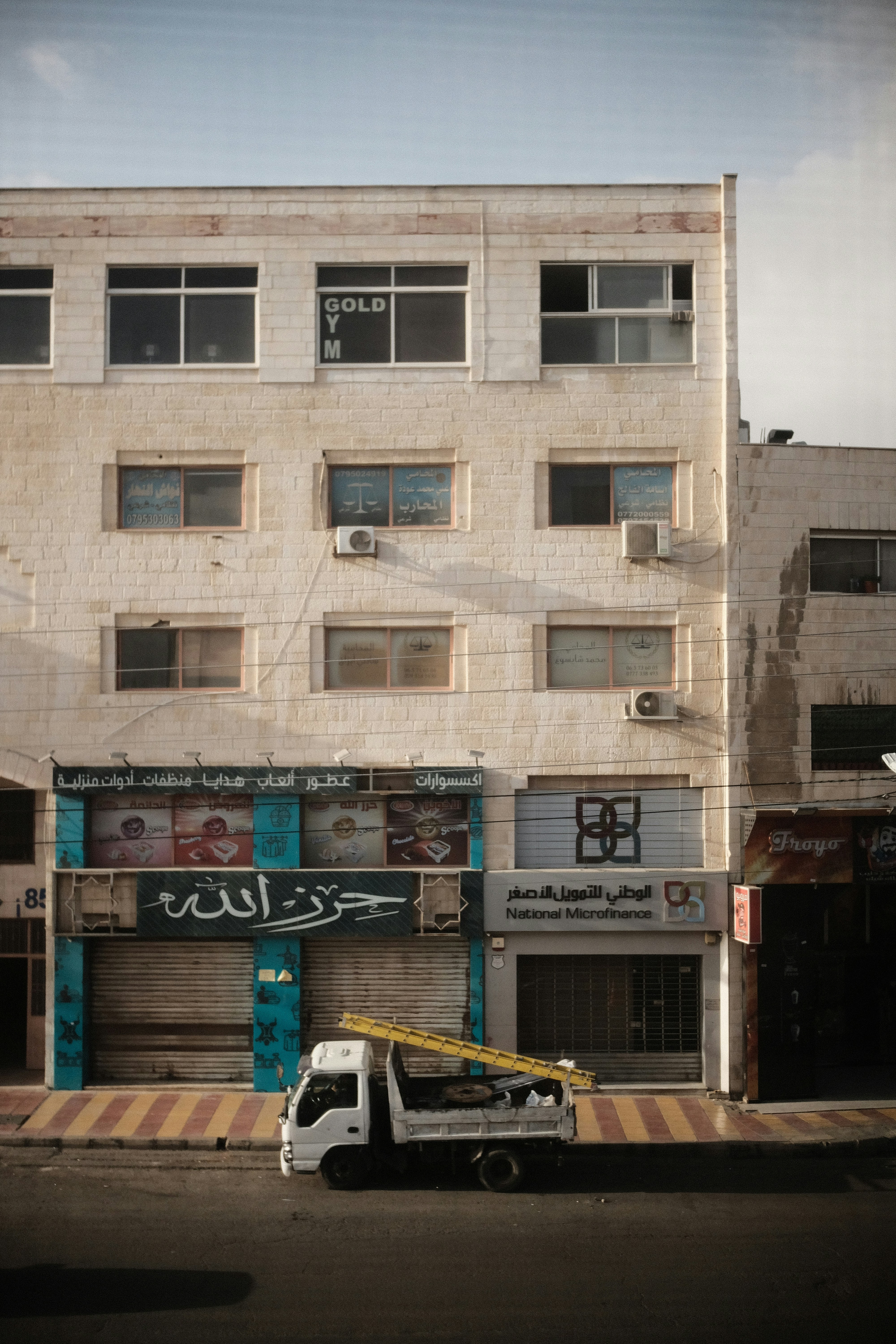 Four-story beige building with a truck parked in front on a deserted street, under a clear sky.