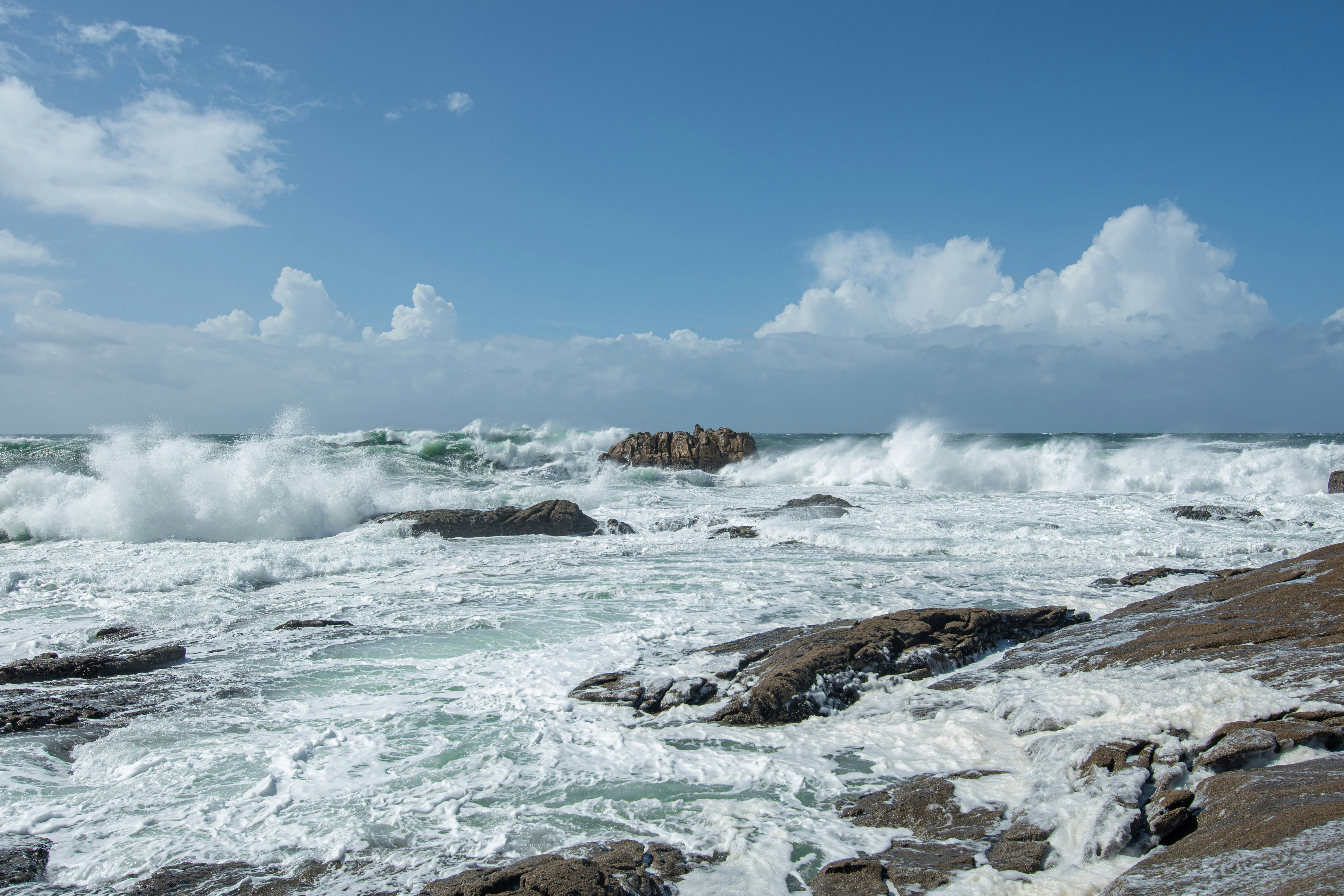 Waves crashing over jagged rocks under a bright blue sky with scattered clouds.