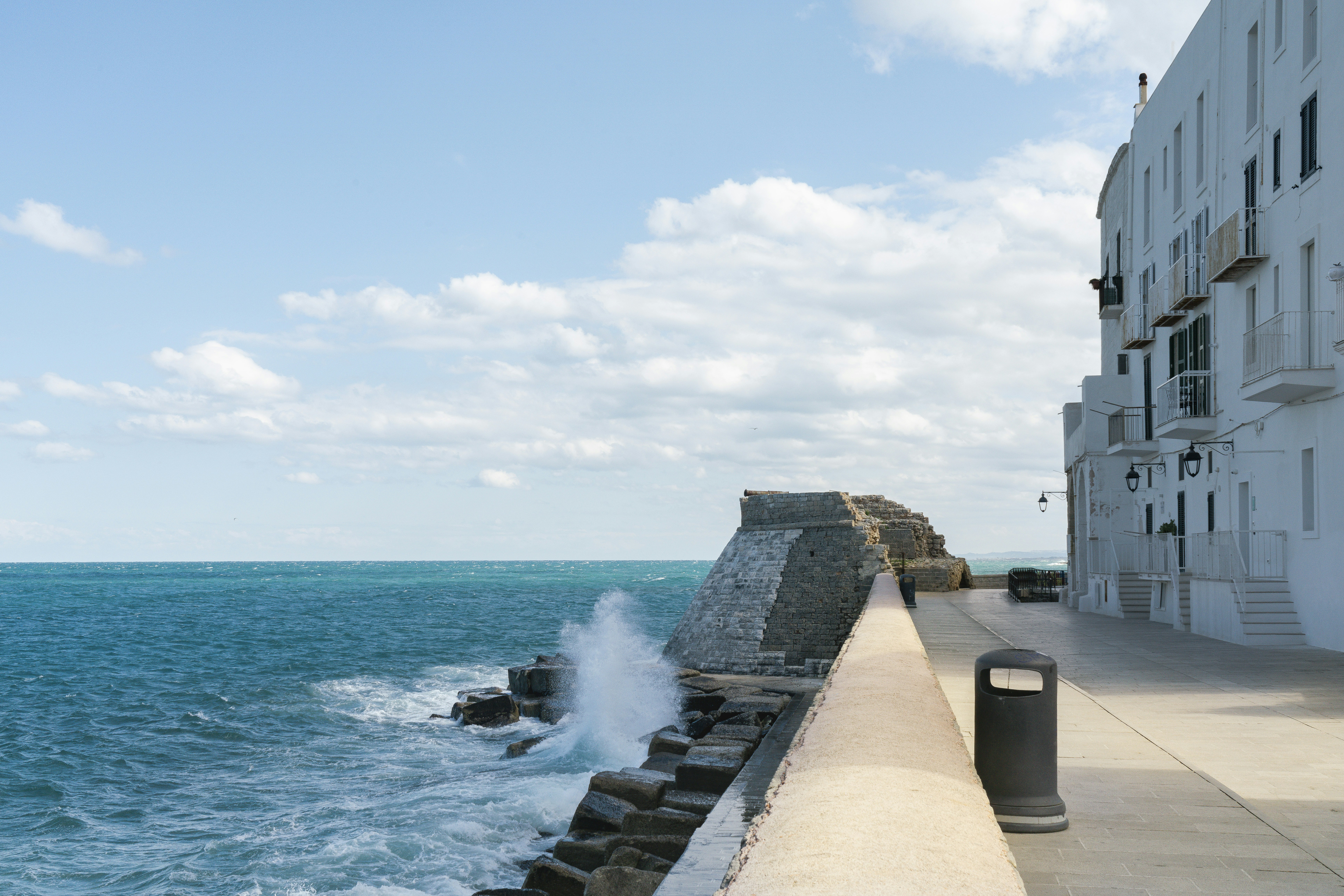 Waves crash against a seawall beside white buildings under a clear blue sky.