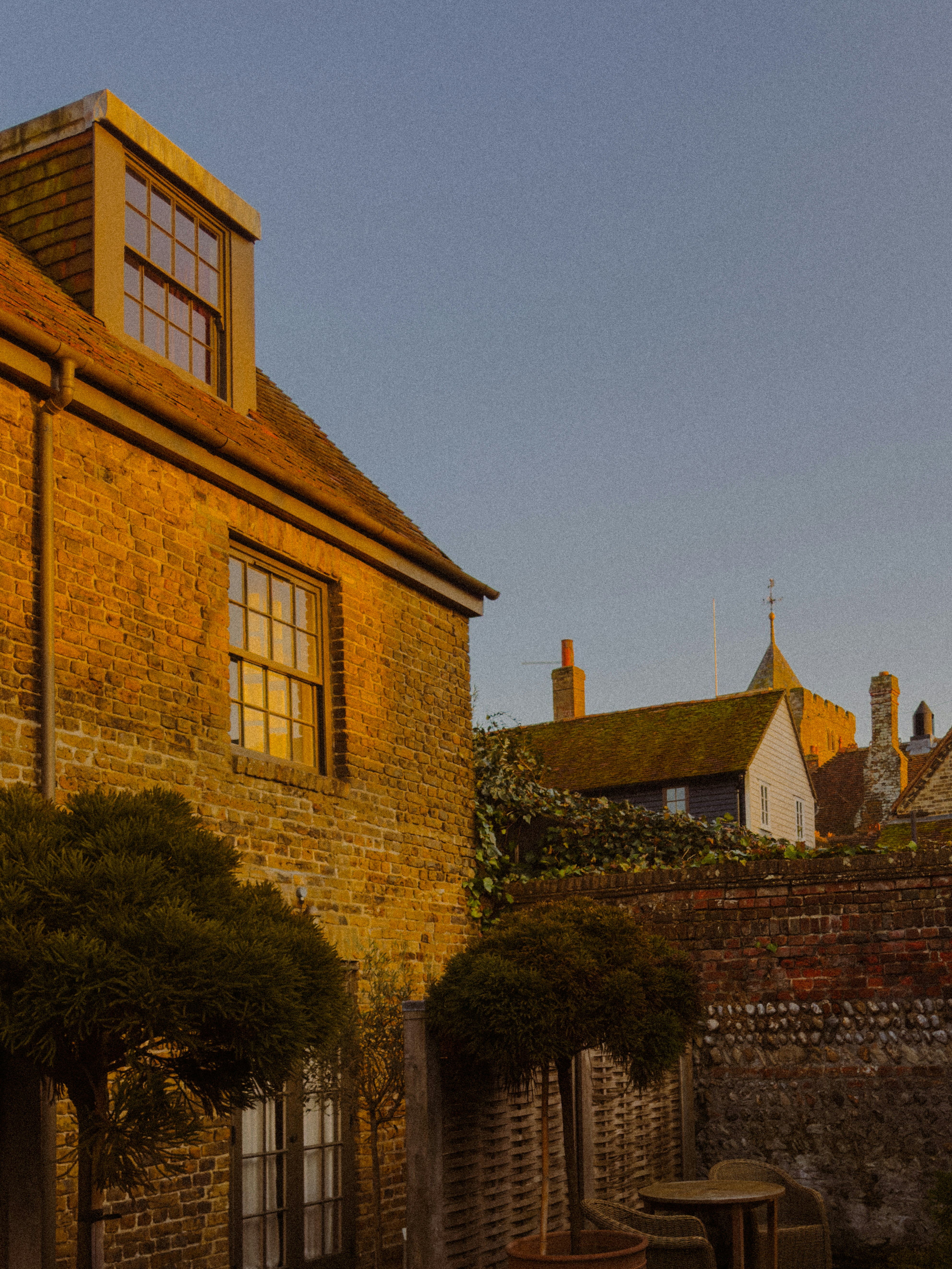 Warm sunlight casts a golden glow on rustic brick buildings and garden walls in a quaint town.