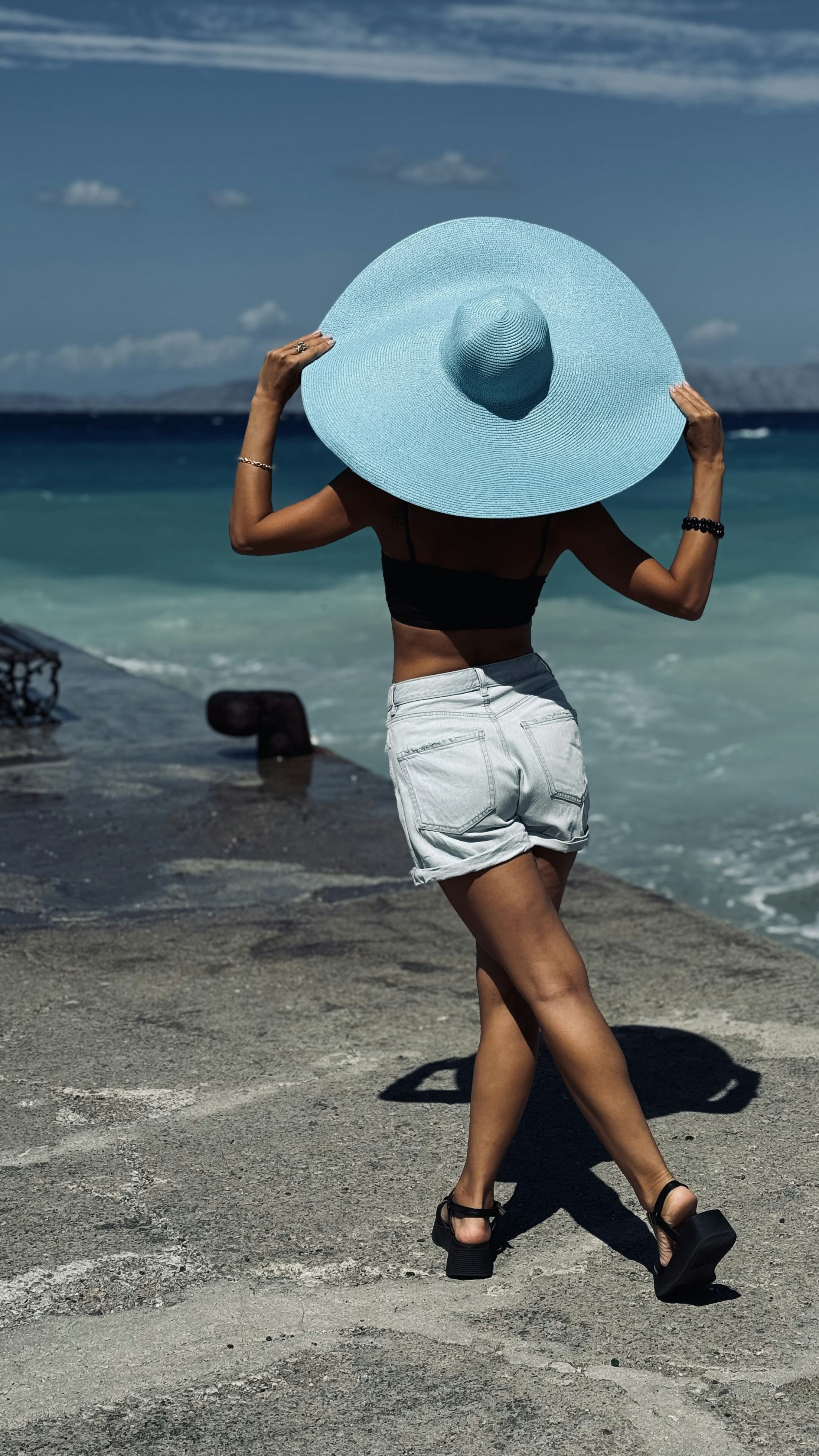 Woman in a wide-brimmed blue hat stands on a seaside pier, facing the ocean under a clear sky.