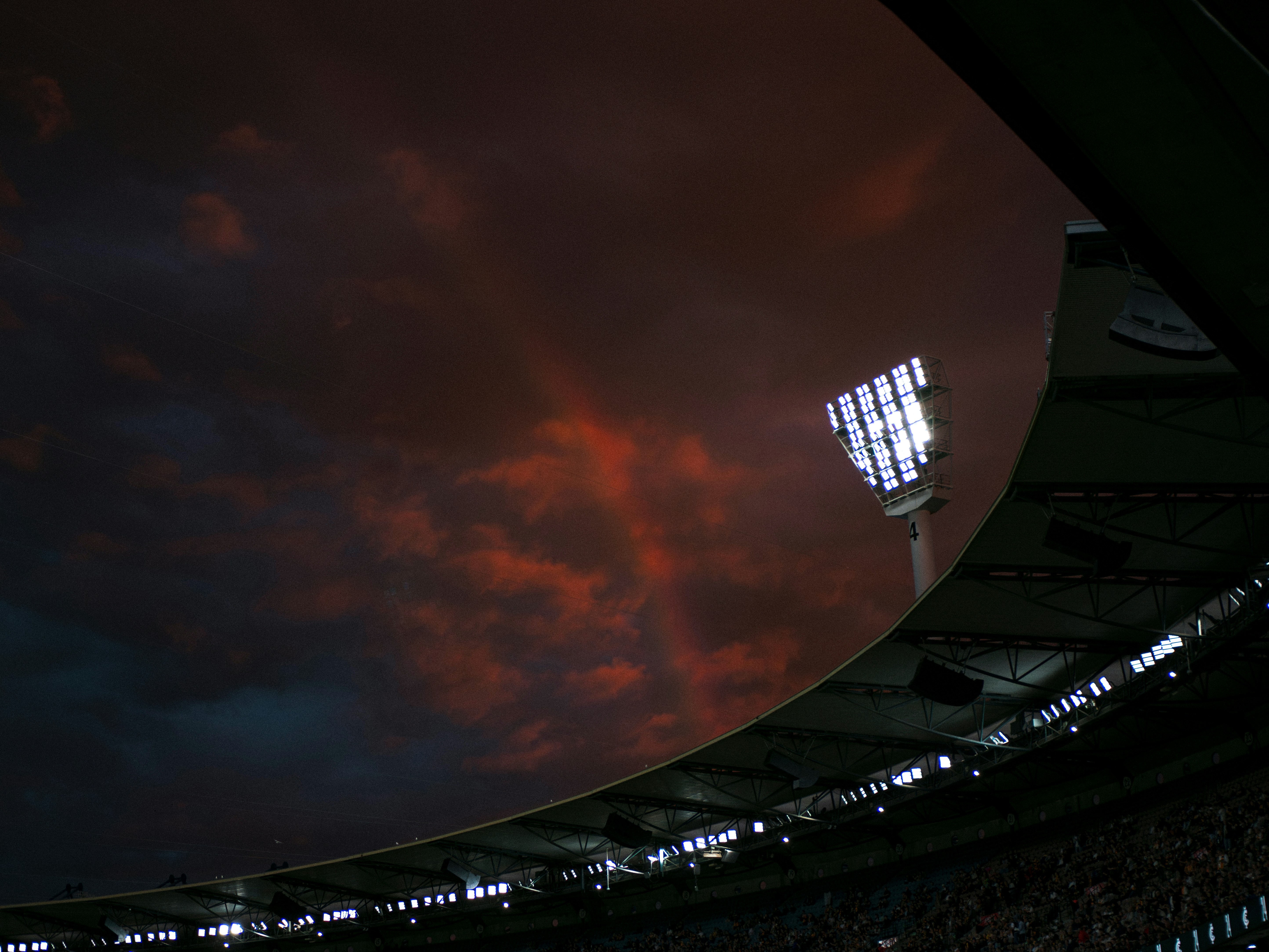 Night shot of a curved stadium roof with bright floodlights piercing the darkness, set against a fiery orange-blue sky.