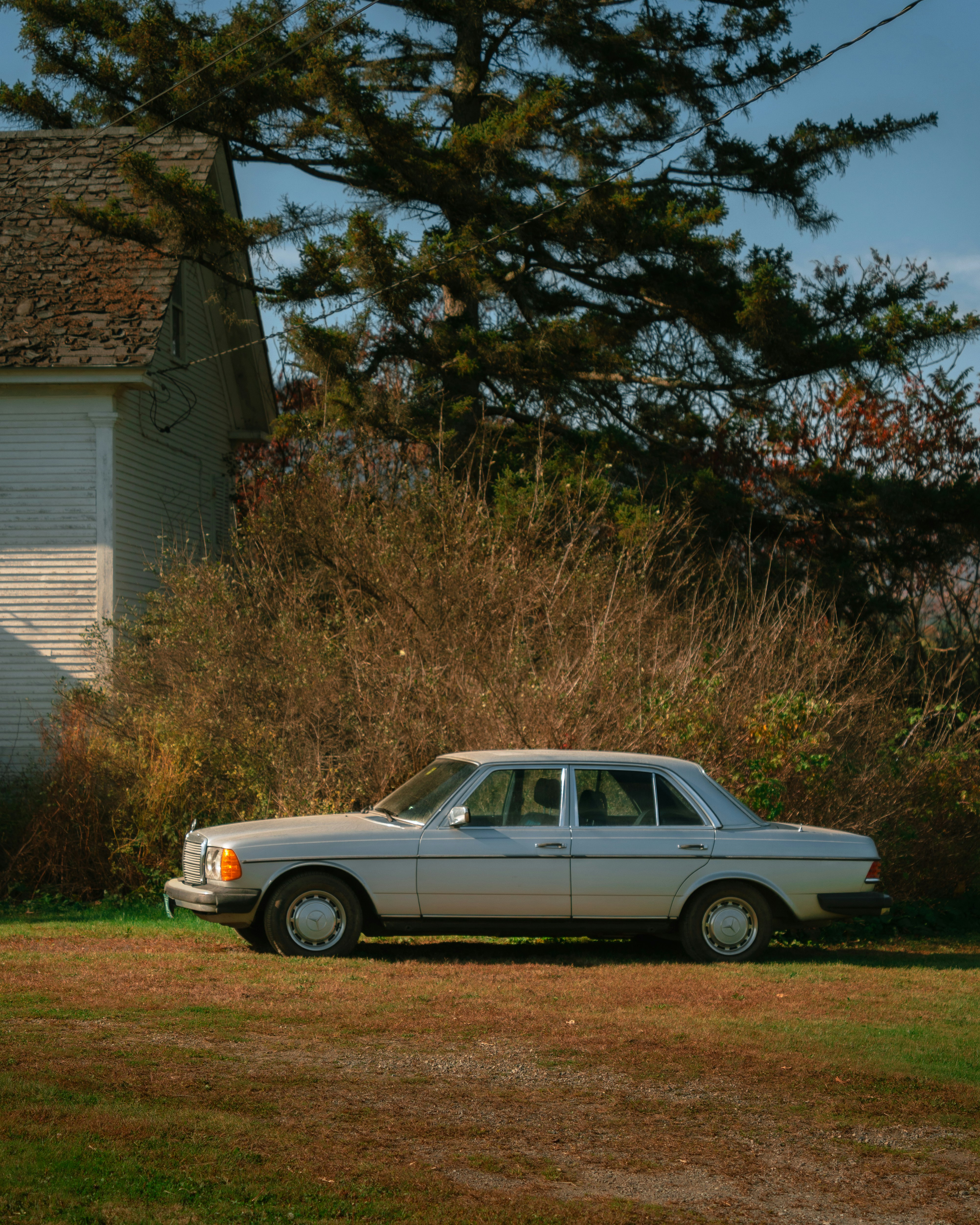 Classic car parked beside an old barn under a clear blue sky, surrounded by autumn foliage.