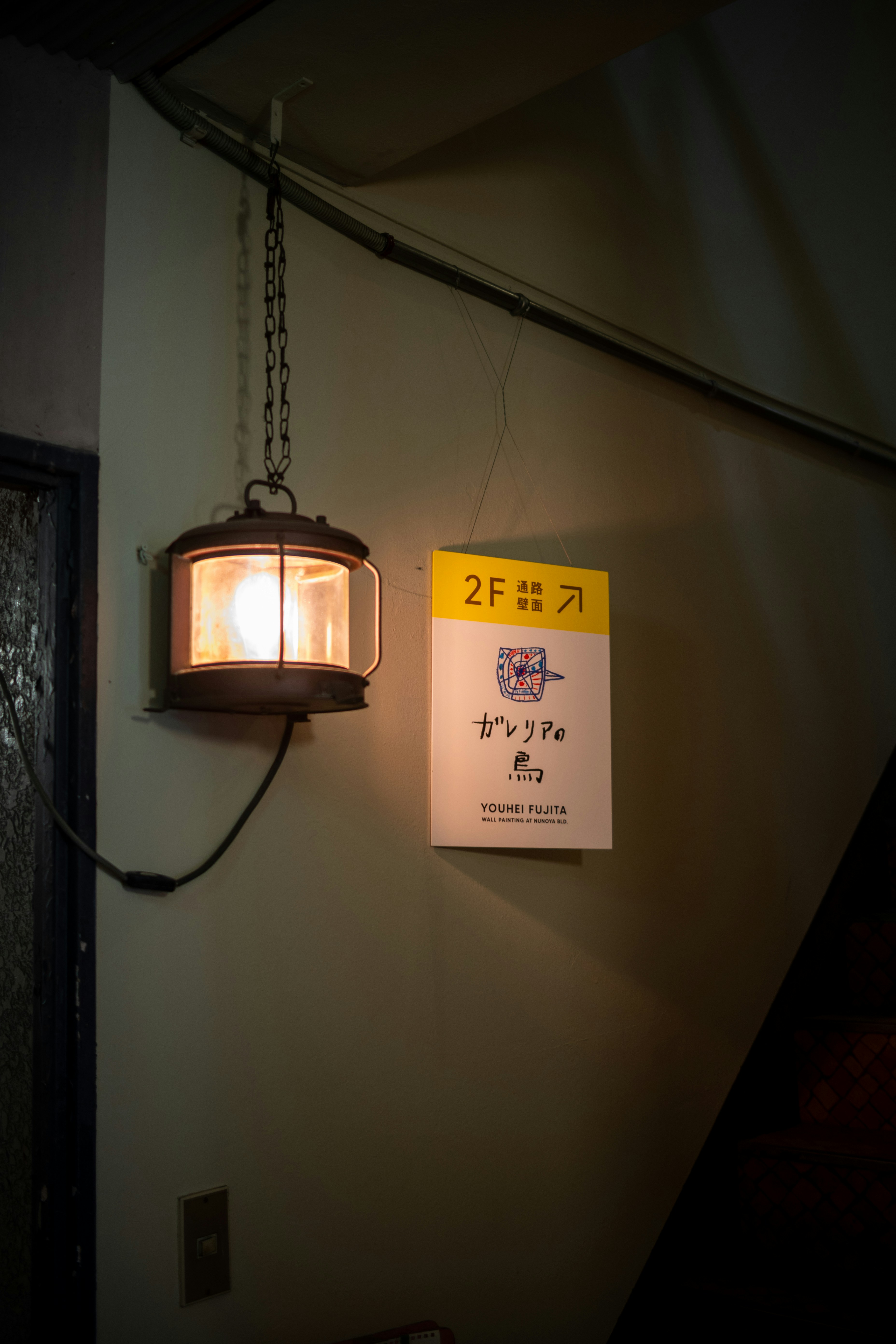 Amber lantern casts a warm glow on a muted stairwell, highlighting a yellow 2F sign mounted on the wall.
