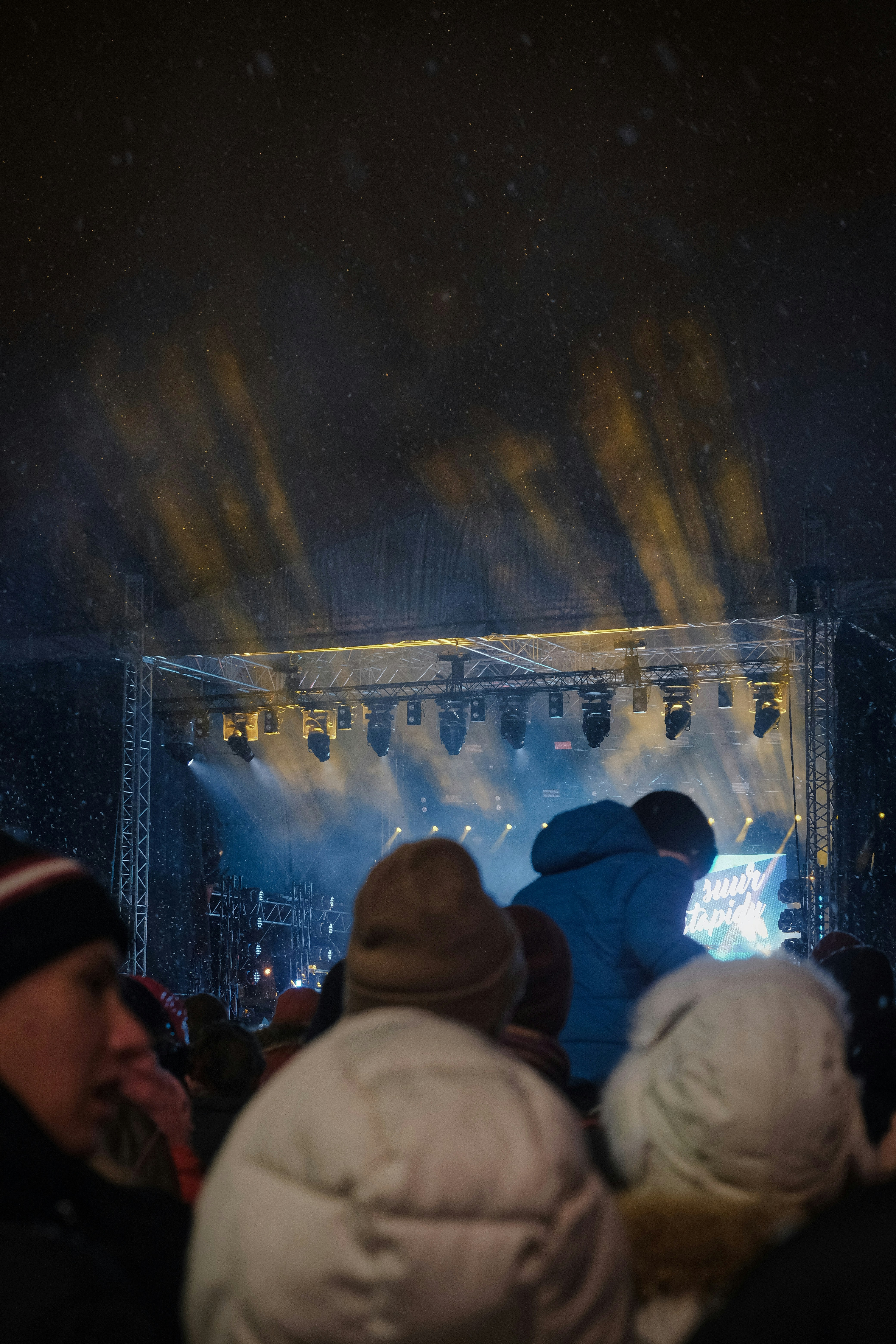 Snowflakes swirl under bright stage lights at a bustling outdoor concert, as attendees bundled in winter attire enjoy the show.