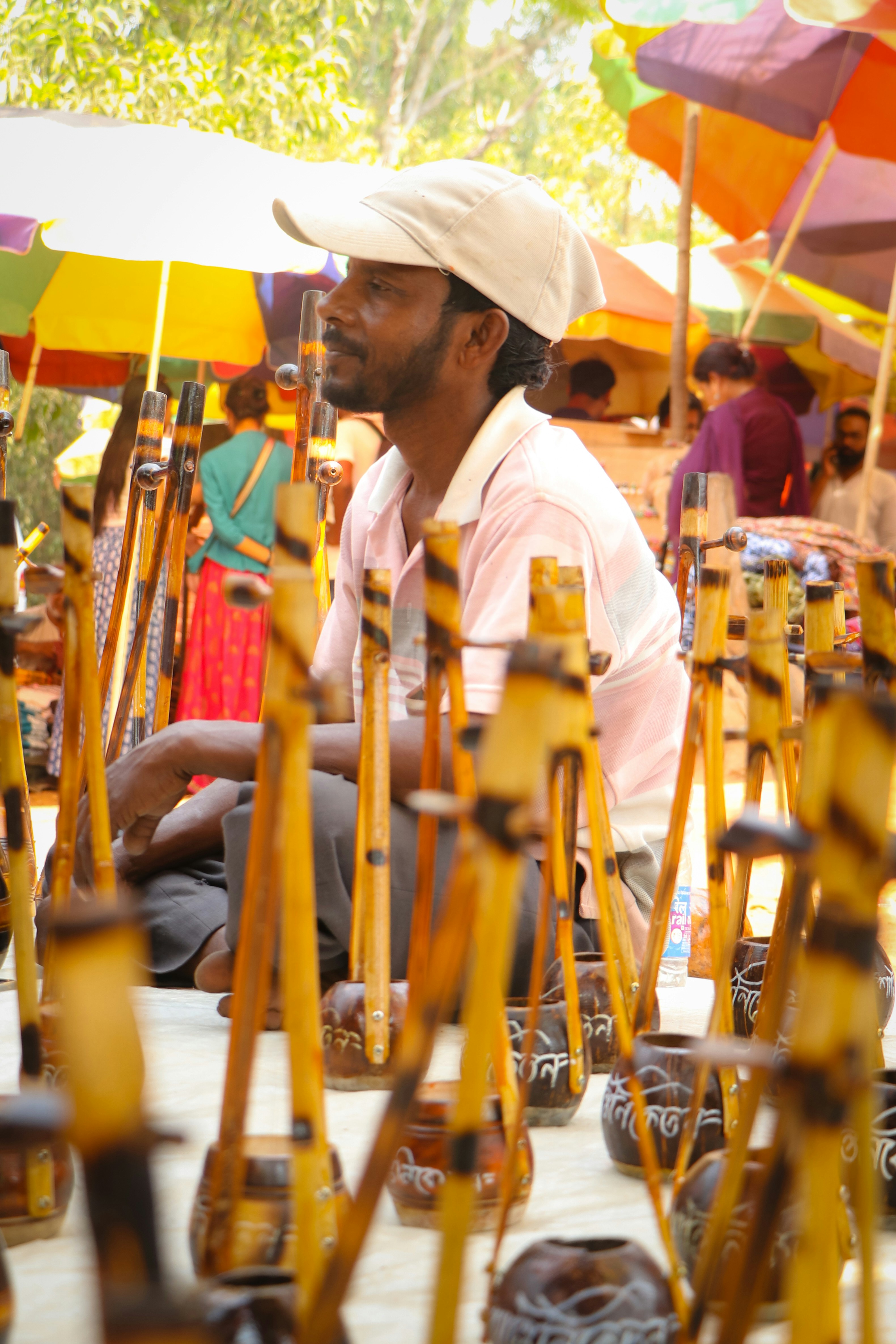 A man in a light pink shirt and beige cap sits among upright string instruments at a sunlit market with colorful umbrellas overhead.