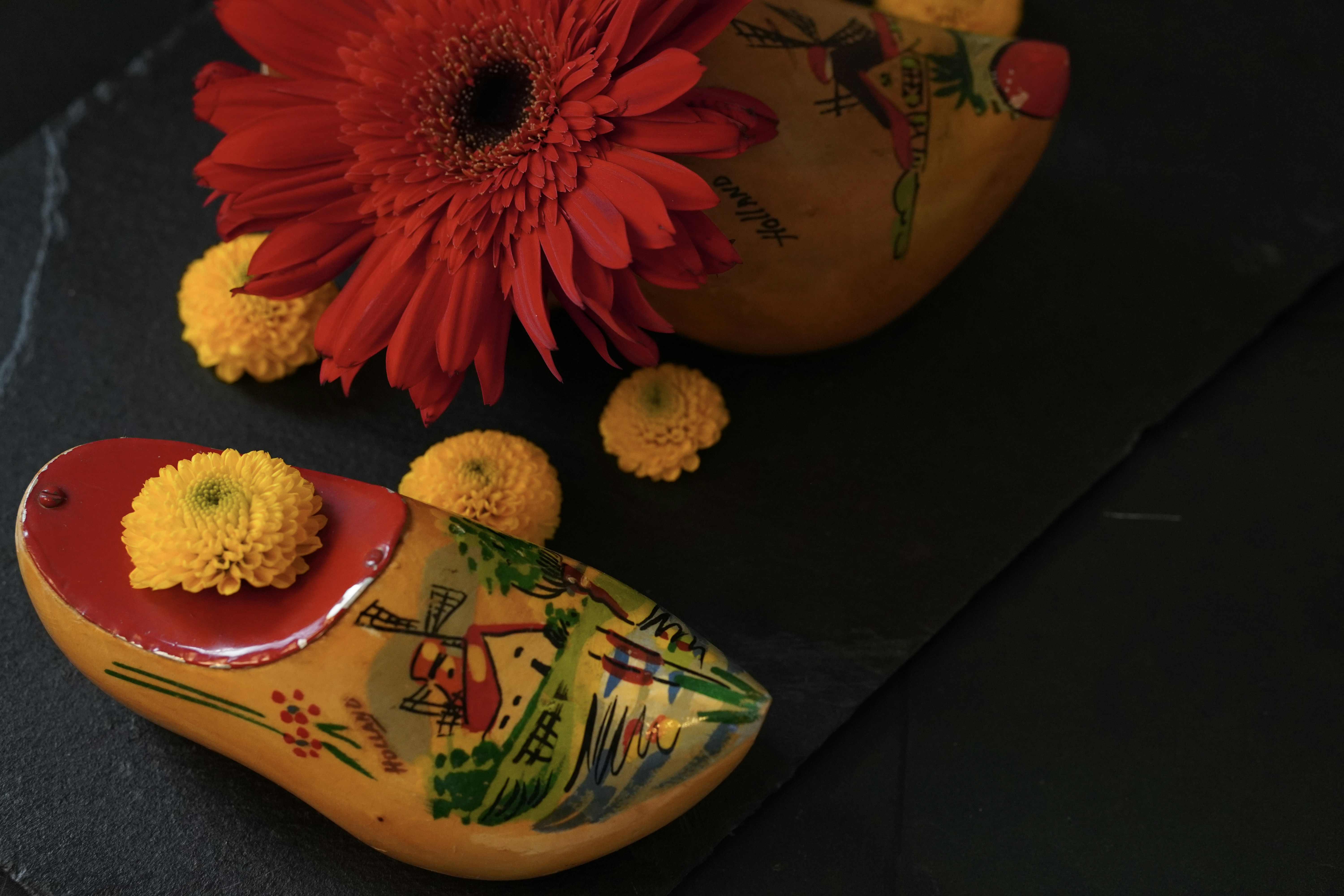 Colorful wooden clogs adorned with floral patterns, accompanied by vibrant gerbera daisies and yellow button flowers on a dark slate background.
