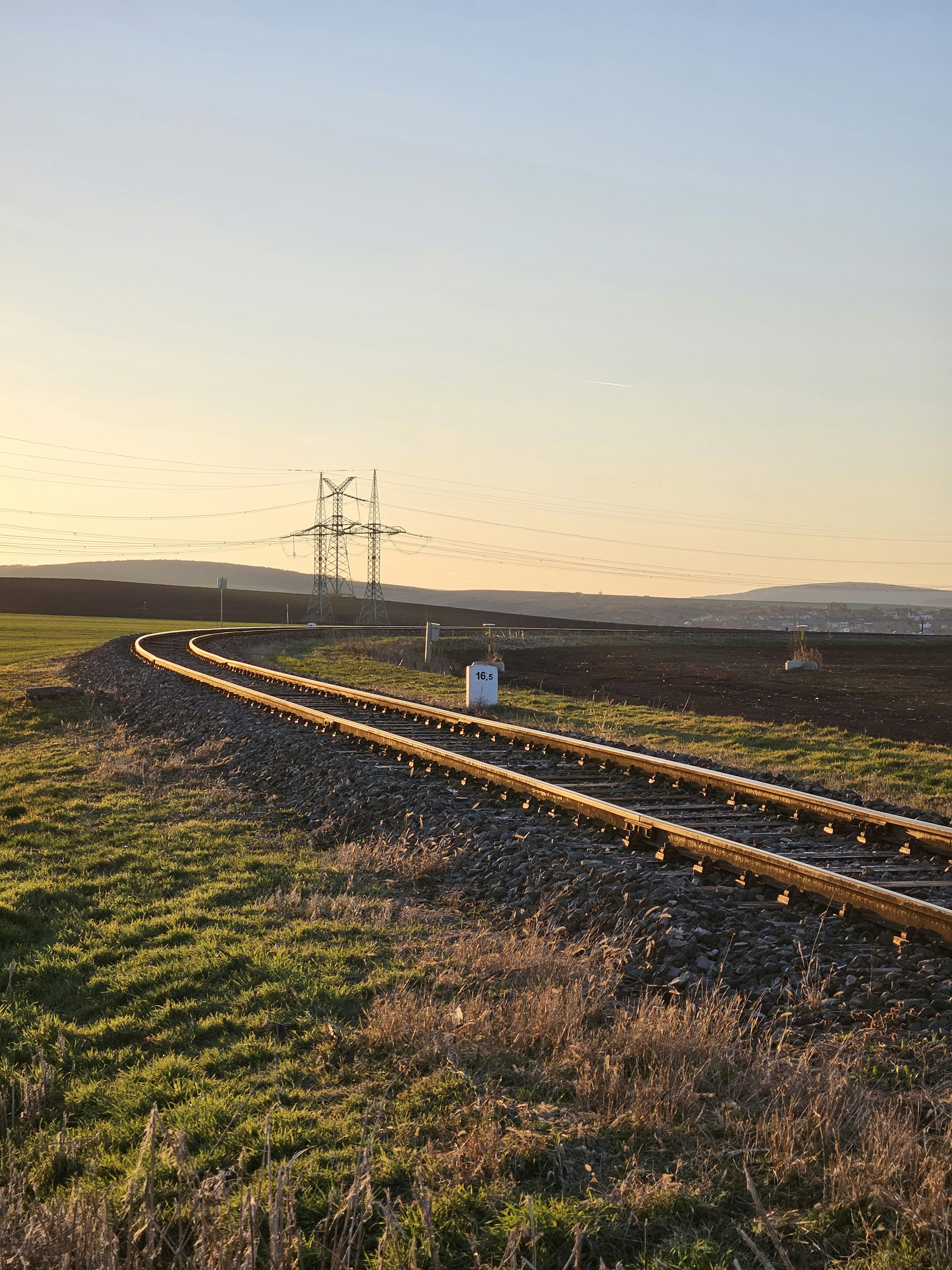 Sunset-lit rural landscape with curved railroad tracks receding toward the distant horizon, flanked by grassy fields. It emphasizes leading lines and the quiet expanse of a rural rail corridor.