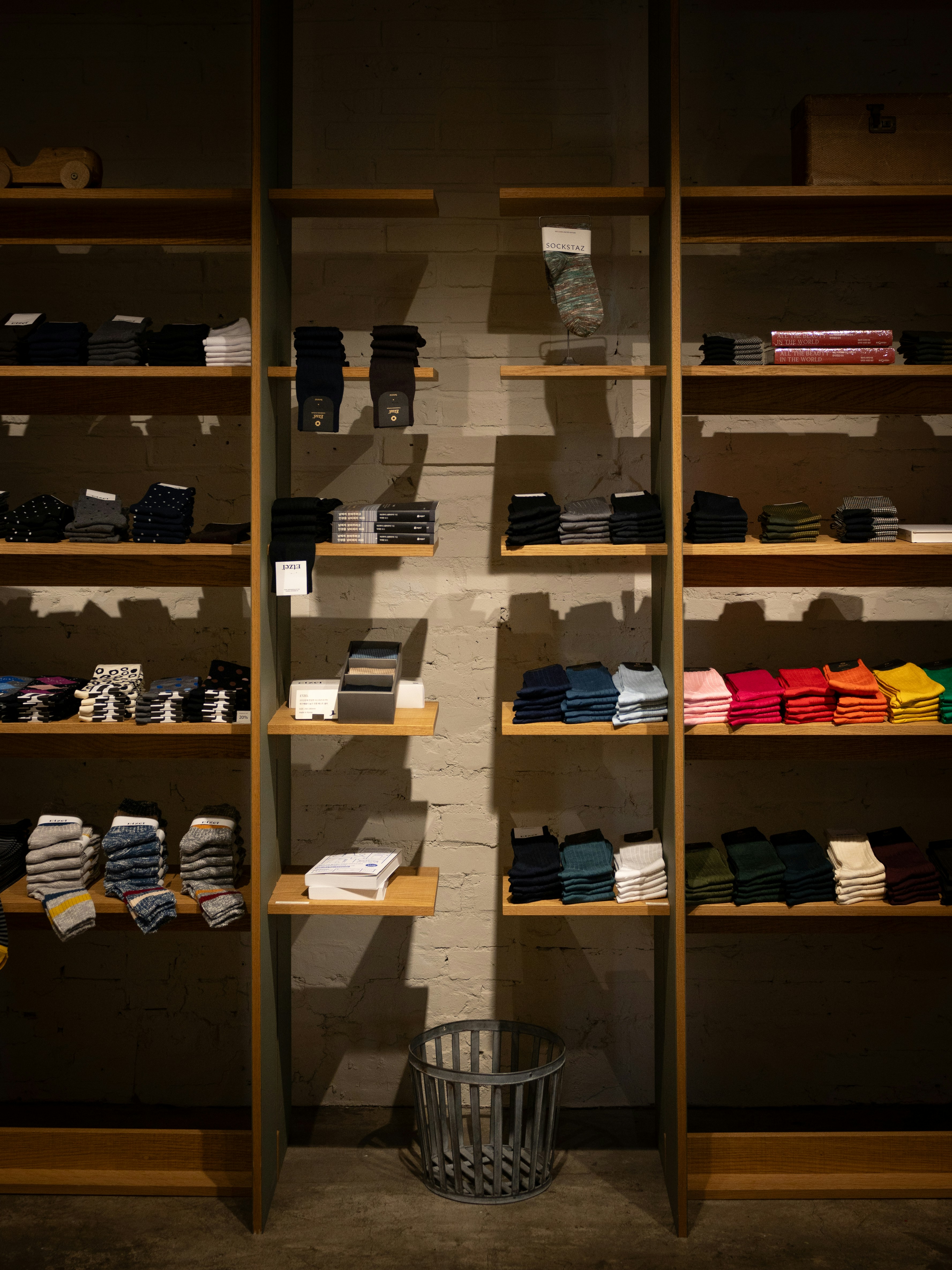 Rows of folded socks on wooden shelves fill a retail wall, with a metal waste basket visible at the bottom center; a moody, warm-toned shop interior. This is a photographed store display.