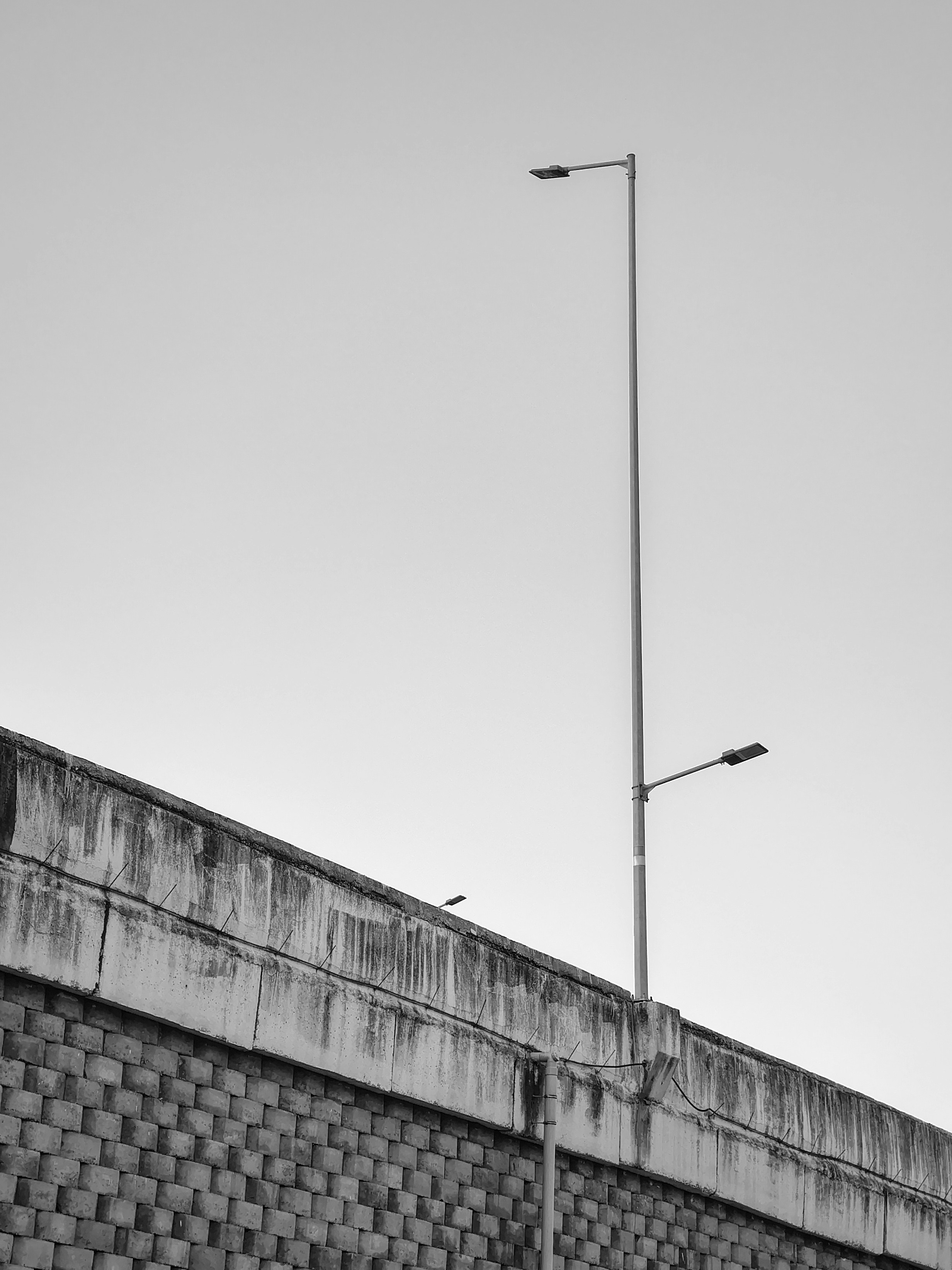 A stark black and white composition featuring a tall streetlight above a textured concrete wall against a clear sky.
