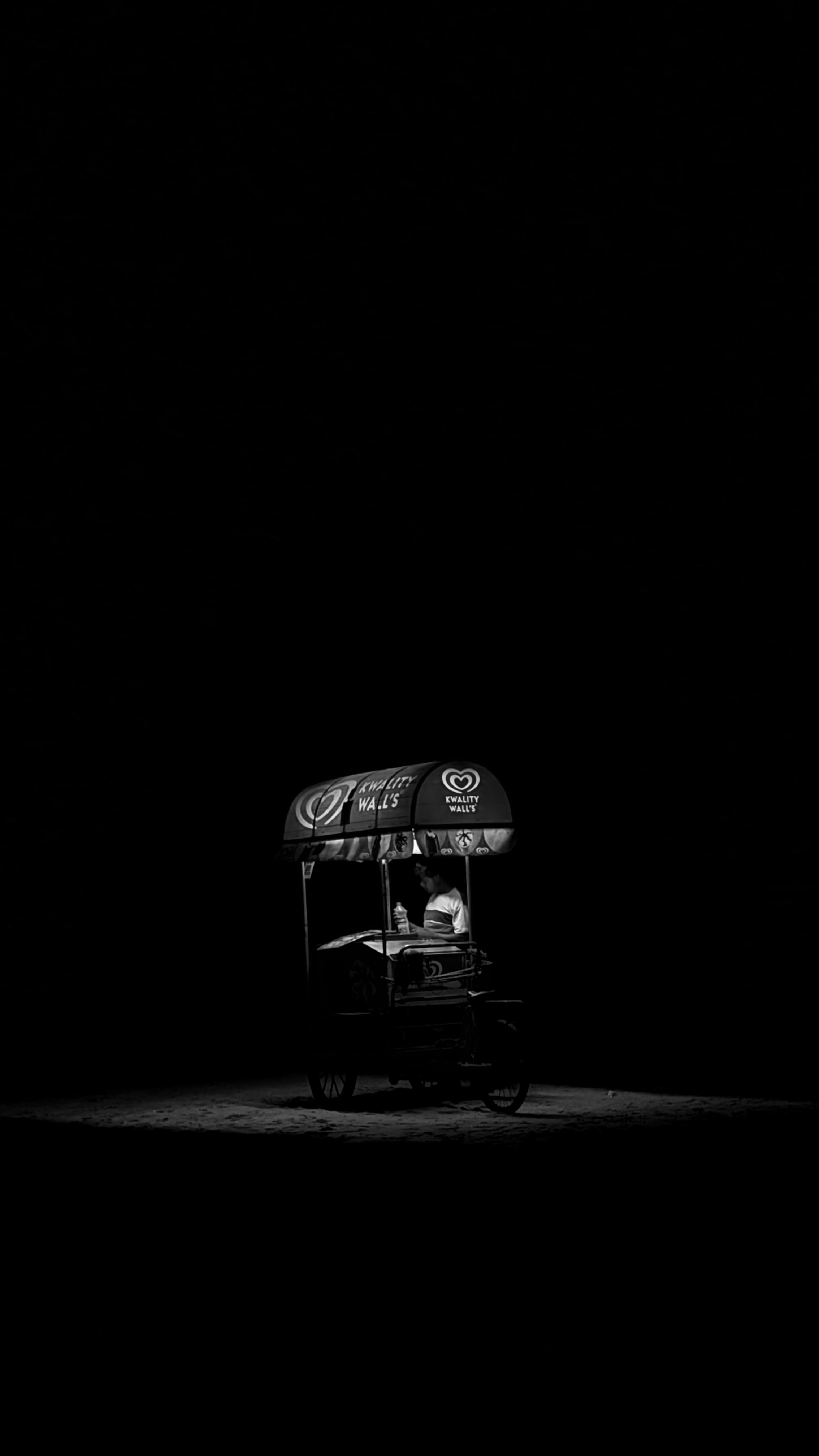 A solitary ice cream vendor sits beneath a dimly lit canopy, surrounded by darkness, highlighting the contrast of light and shadow.