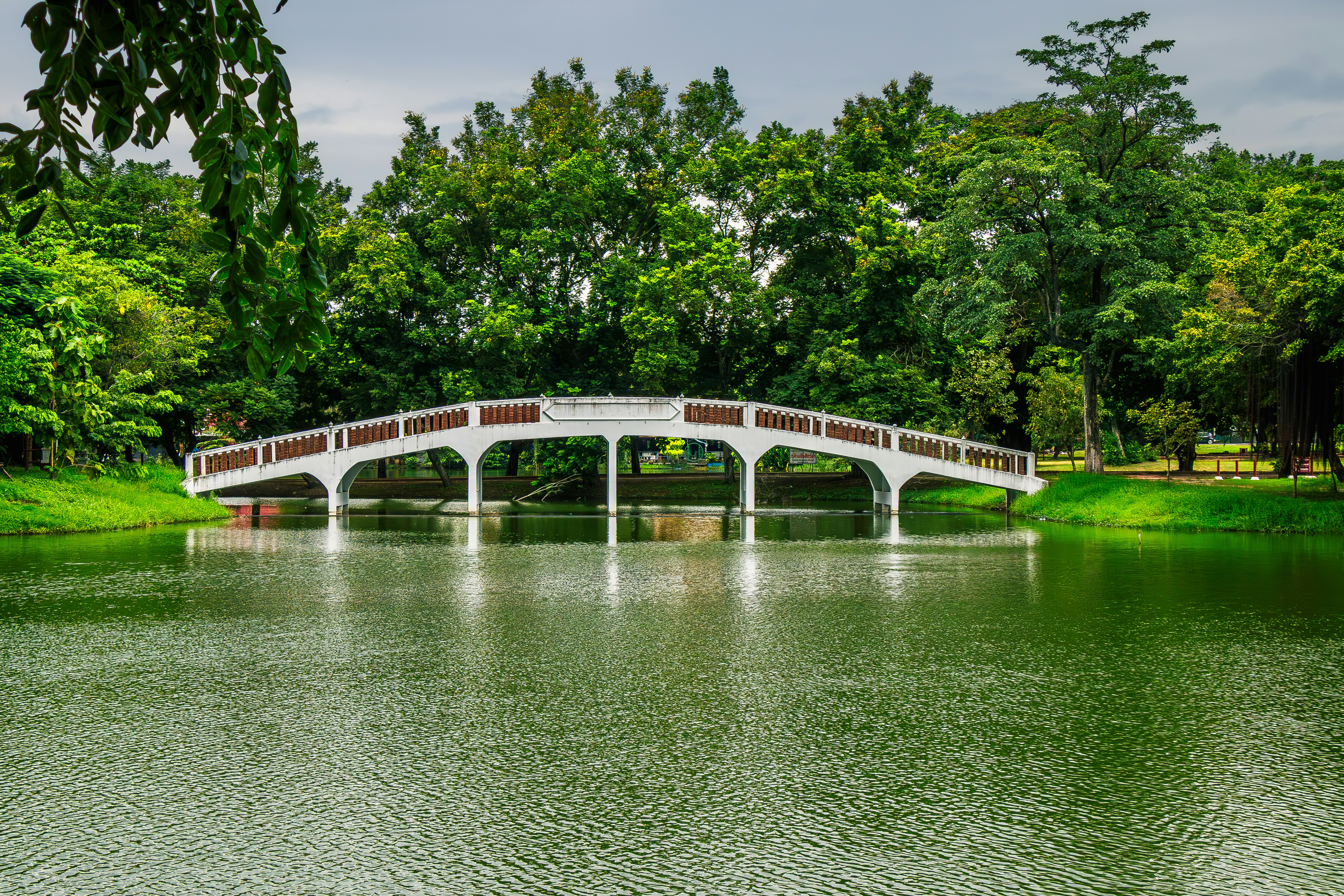 White arched bridge spanning a calm, tree-lined pond under an overcast sky.