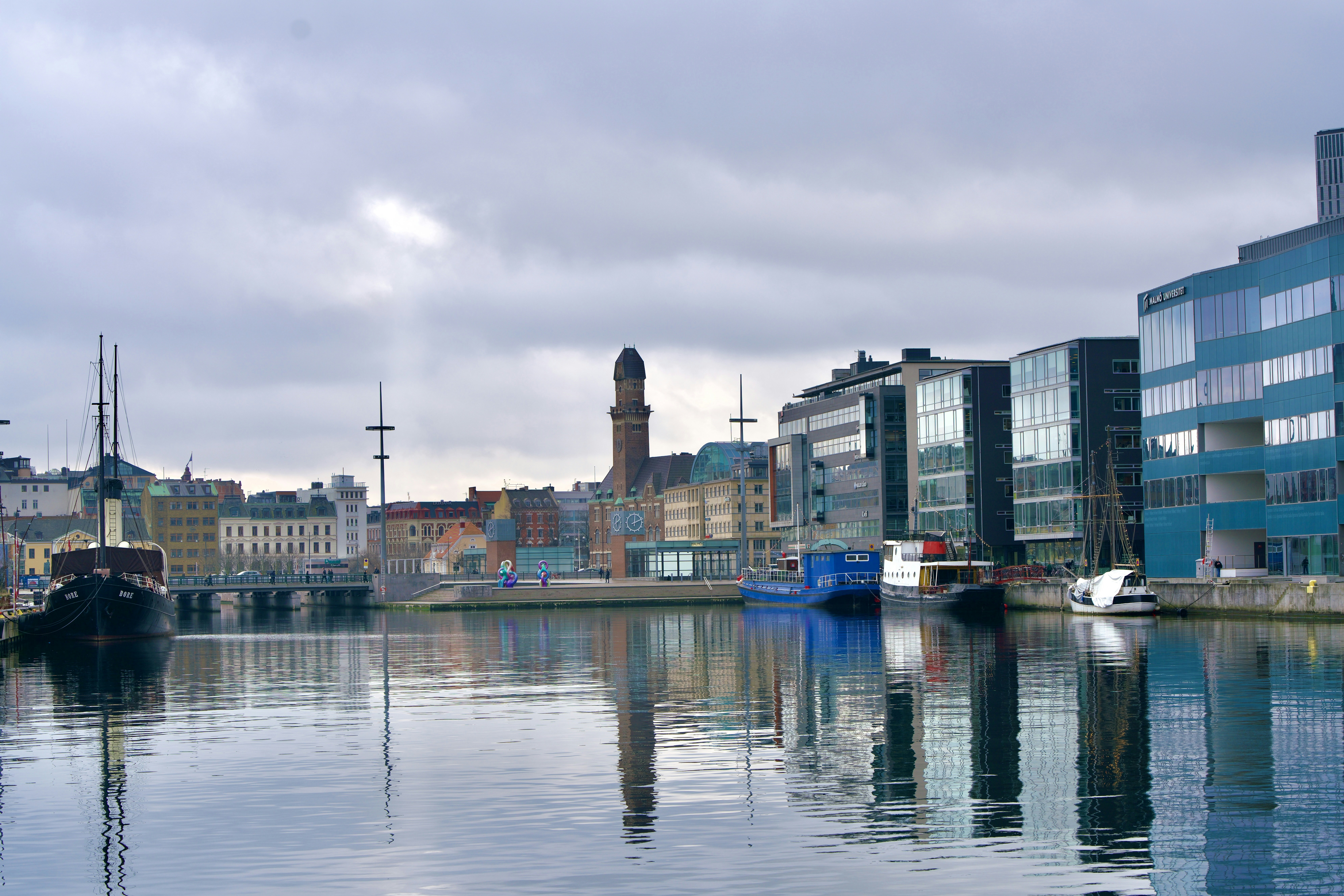 Modern buildings and boats reflected in a calm harbor under a cloudy sky.