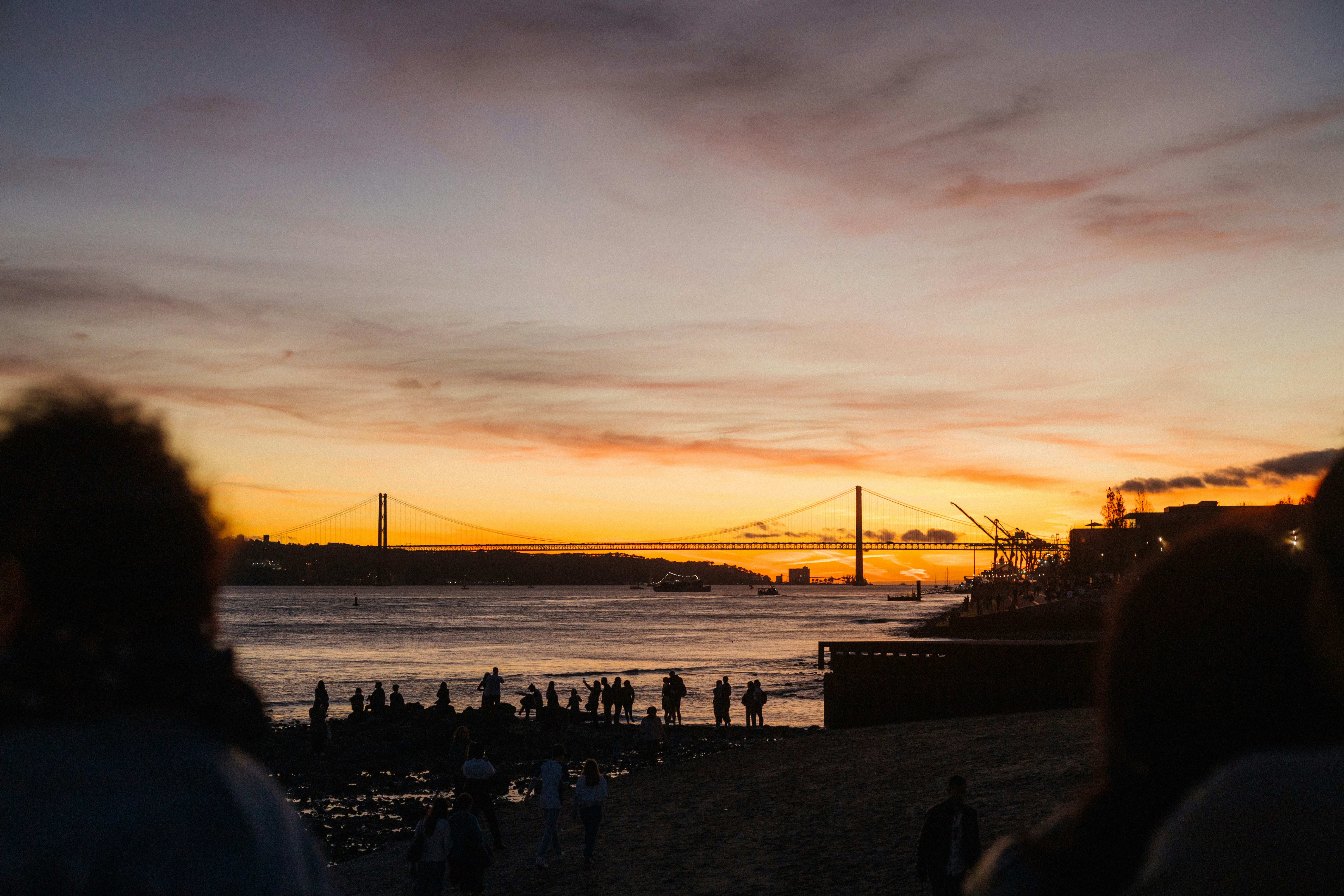 Silhouetted figures gather by the waterfront as the sun sets behind the 25 April Bridge in Lisbon.