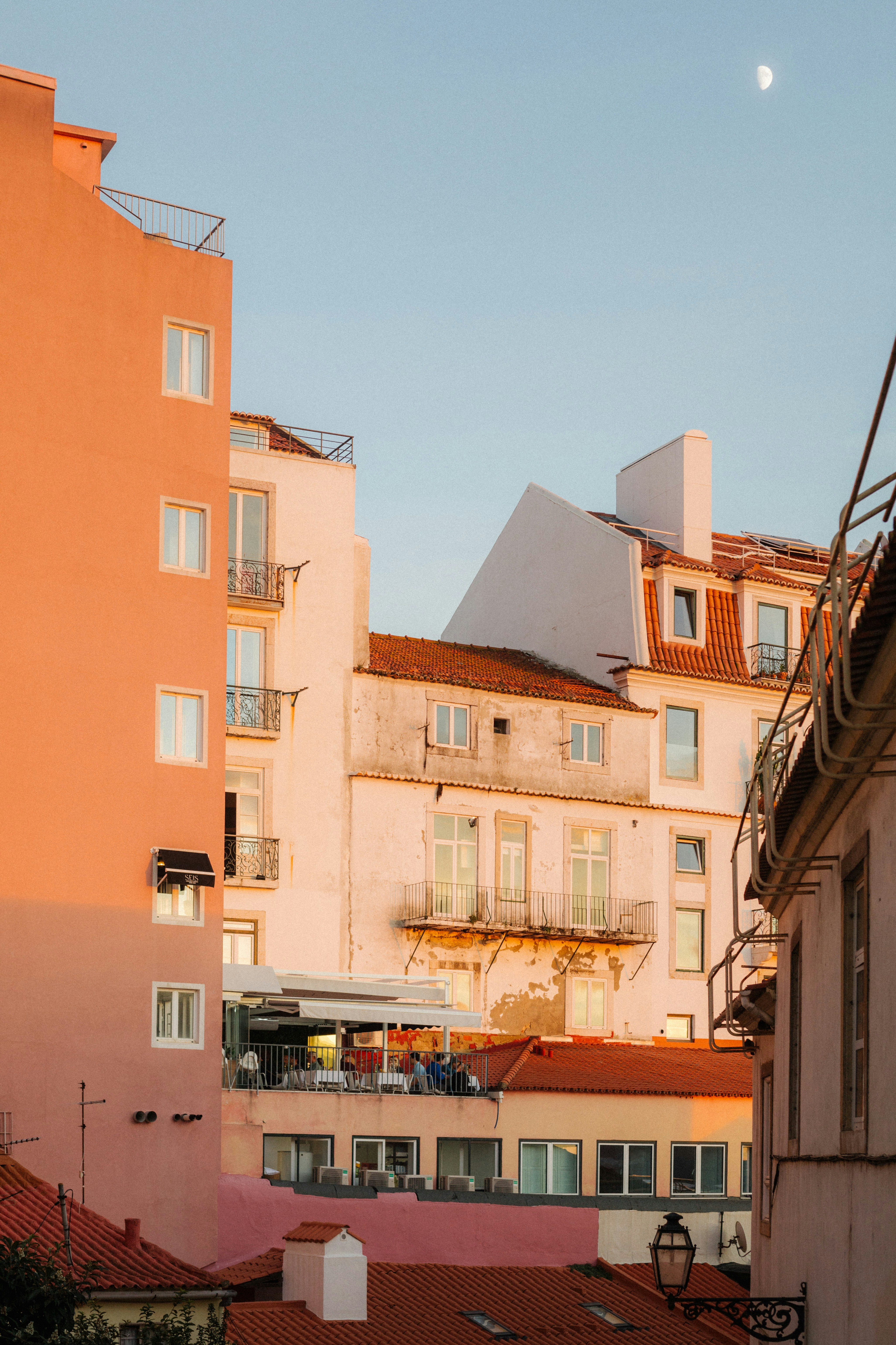 Warm sunlight casts a golden hue on city buildings with a faint moon visible in the clear sky.