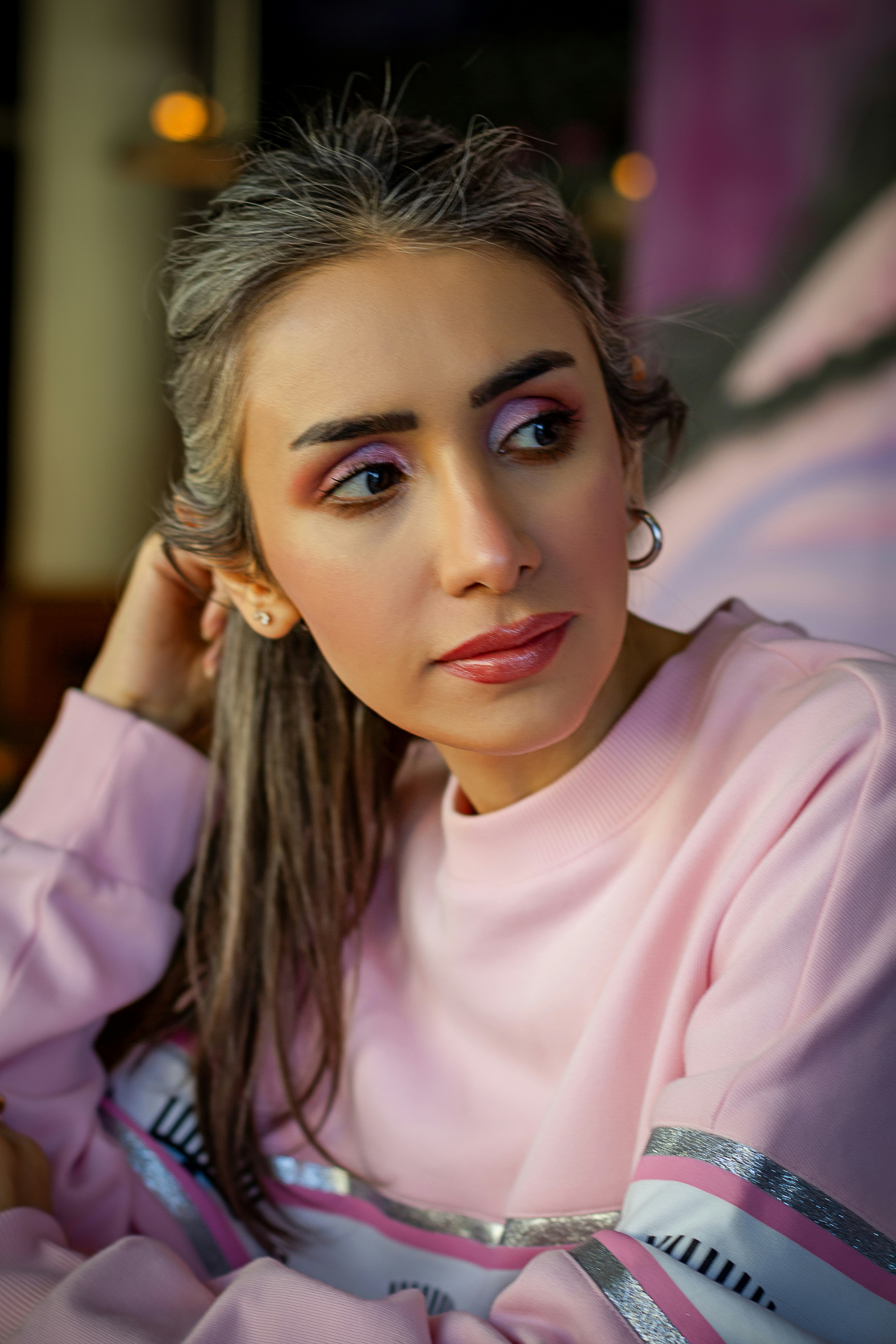 A young woman with striking makeup gazes thoughtfully, dressed in a pastel pink sweater. The soft background hints at a cozy atmosphere.