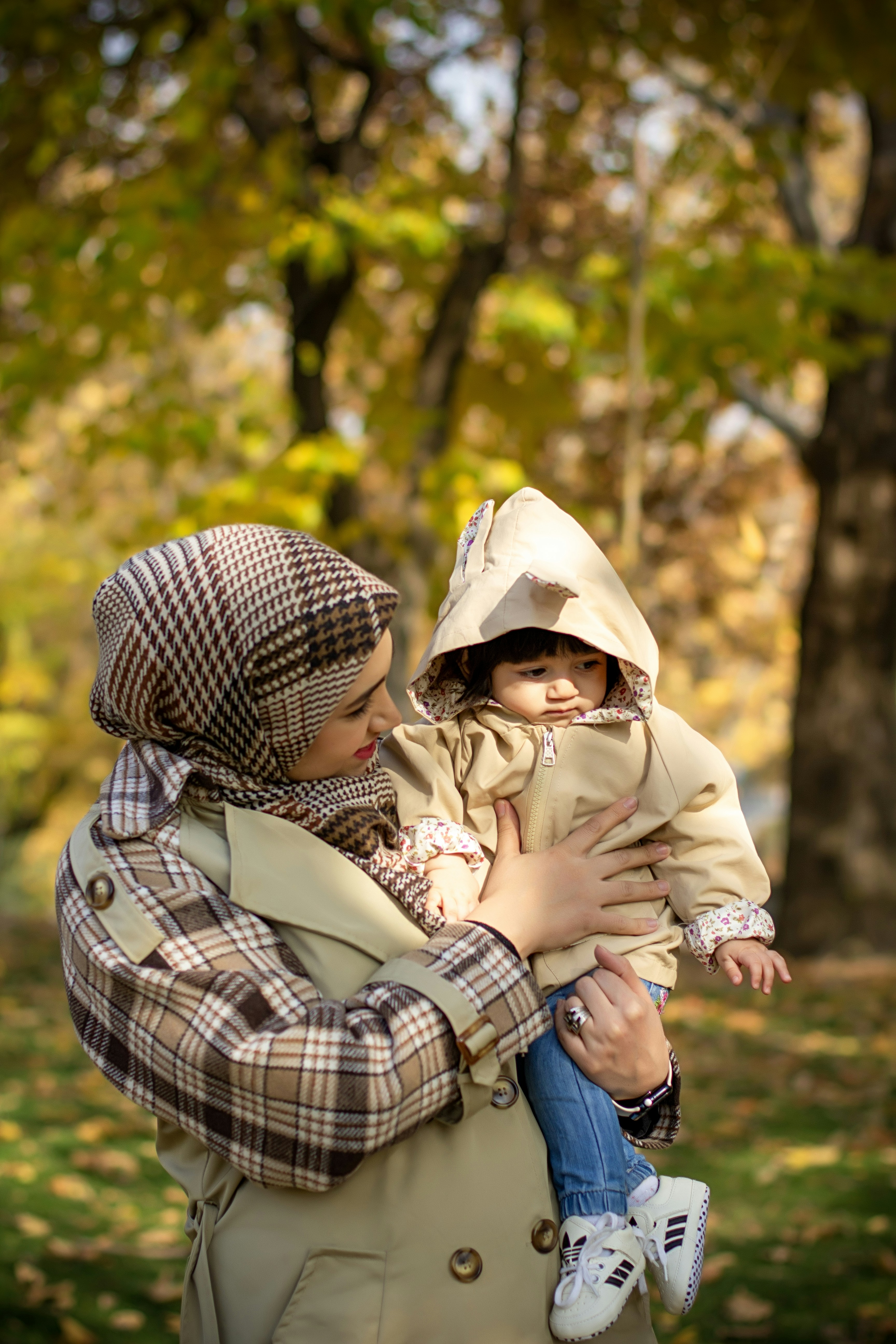 A mother gently holds her child in a cozy beige outfit, surrounded by vibrant autumn foliage. The warmth of their bond contrasts beautifully with the seasonal backdrop.