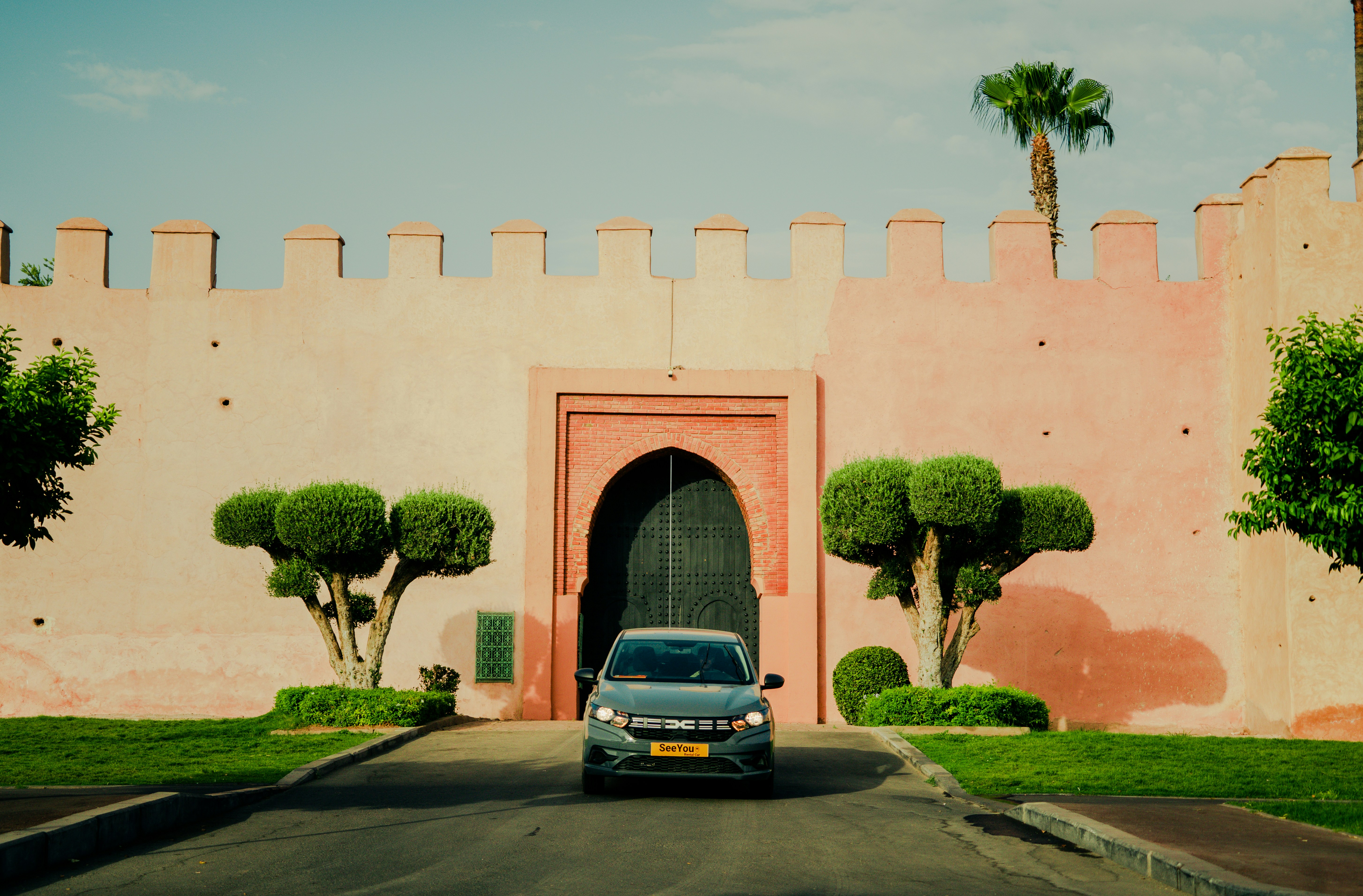Car parked in front of a large, pink fortress wall with manicured trees and a clear blue sky.