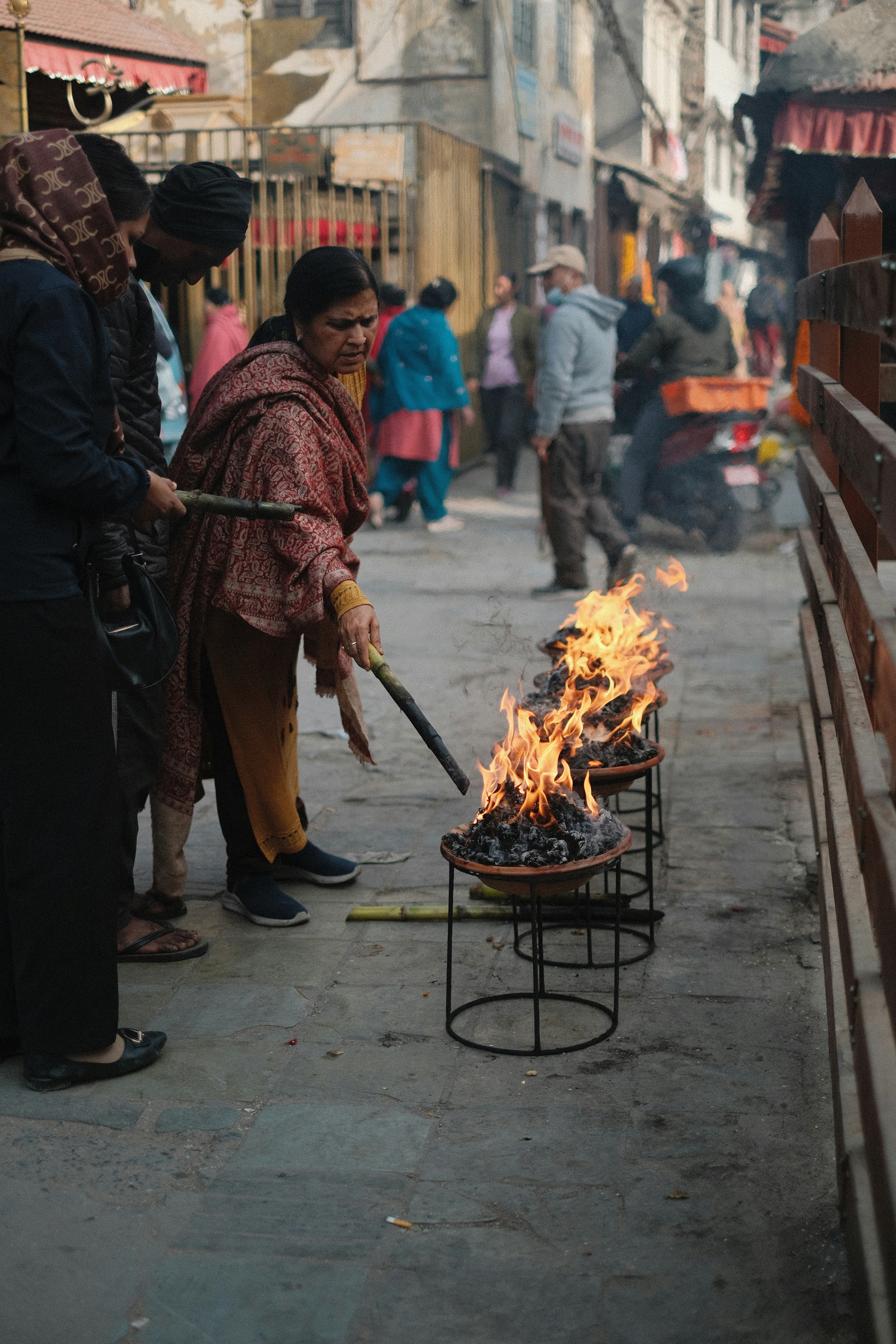 Ember on the Lane: a woman tends street braziers as a bustling market passes by