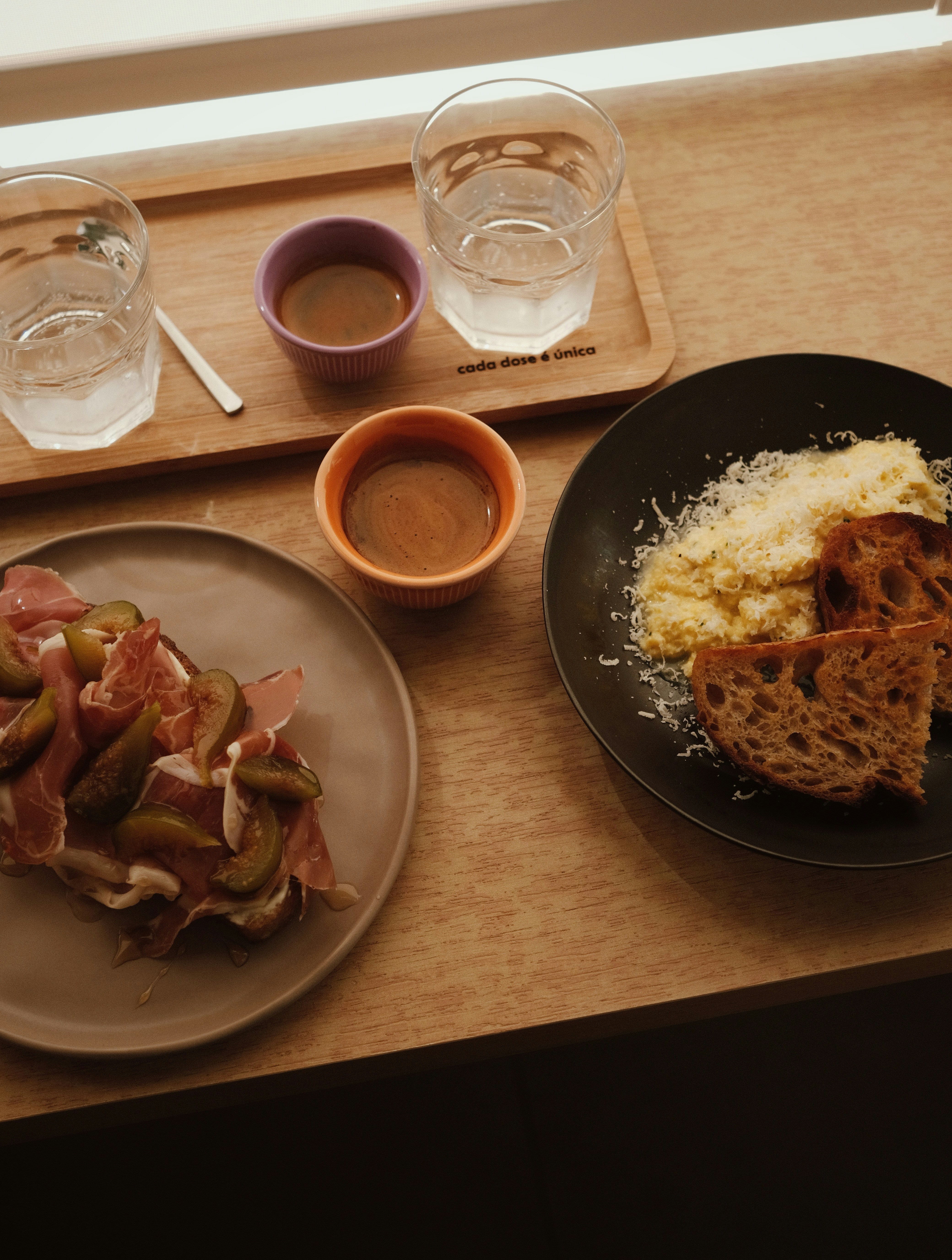Plate featuring sliced prosciutto and pickles alongside scrambled eggs, artisanal bread, and colorful dipping sauces. Wooden tray complements the rustic presentation.