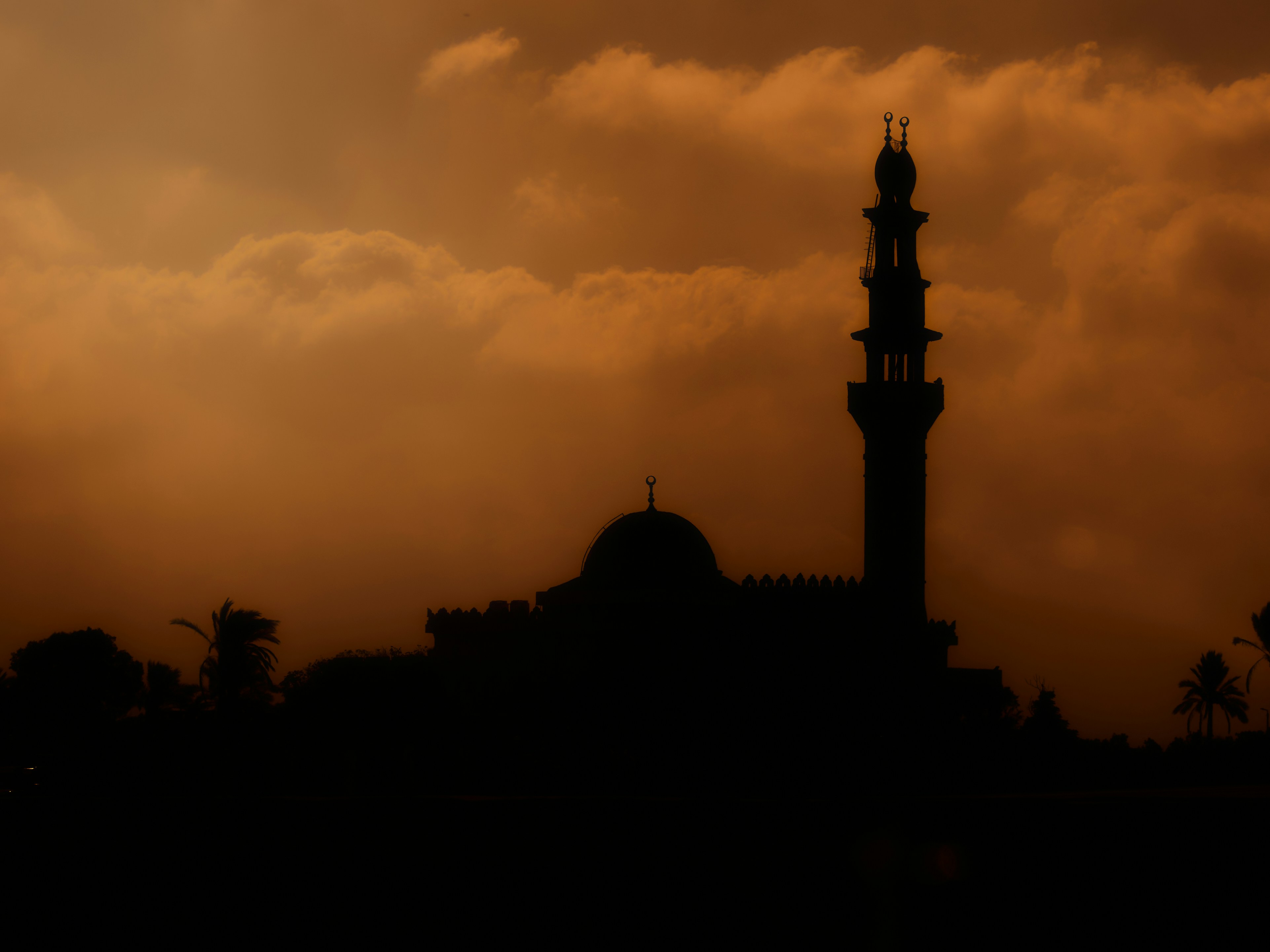 Mosque silhouette with a minaret and dome against a dramatic sunset sky, framed by palm trees.