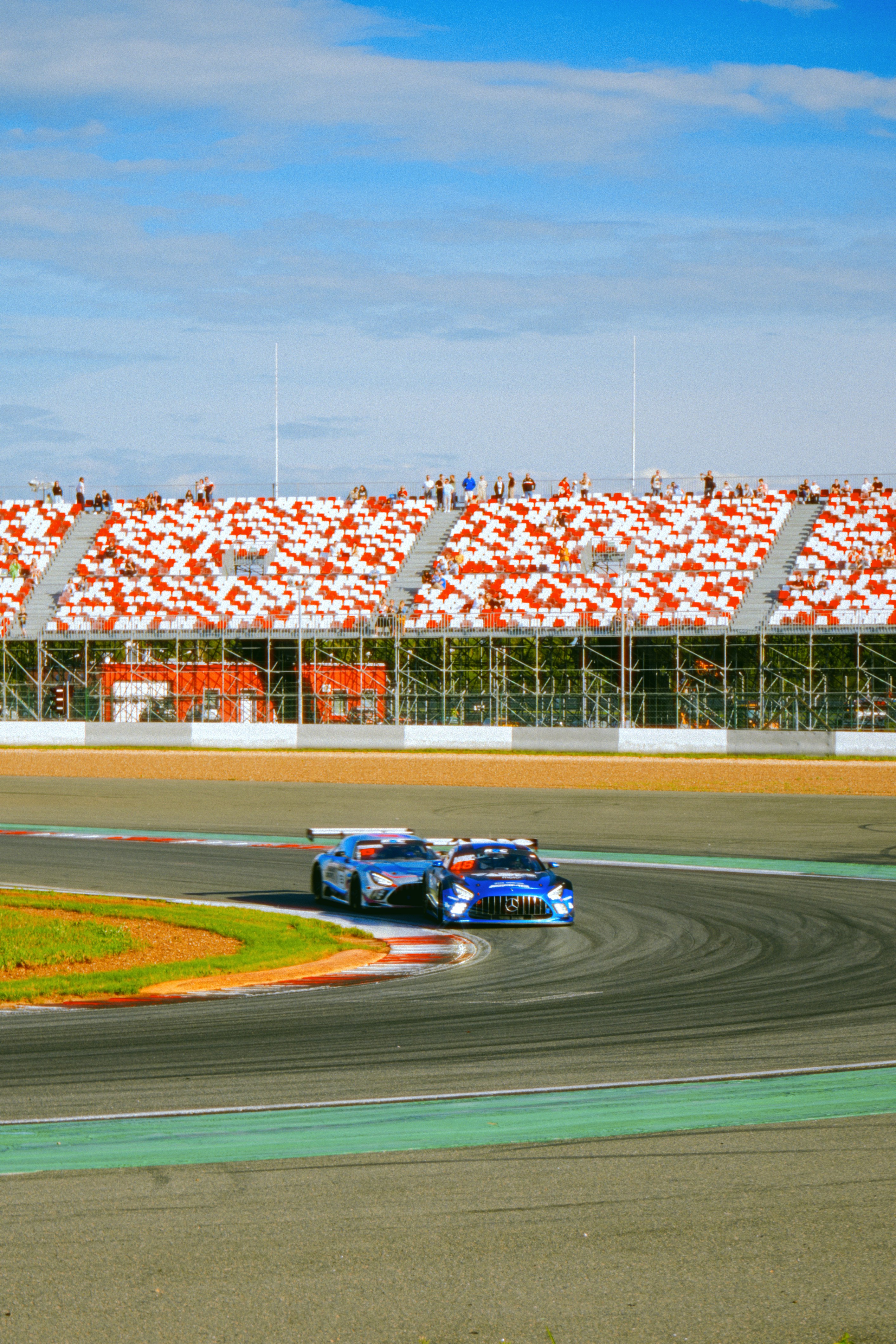Two blue race cars navigate a curve on a sunlit track with a stadium filled with red and white seats in the background.