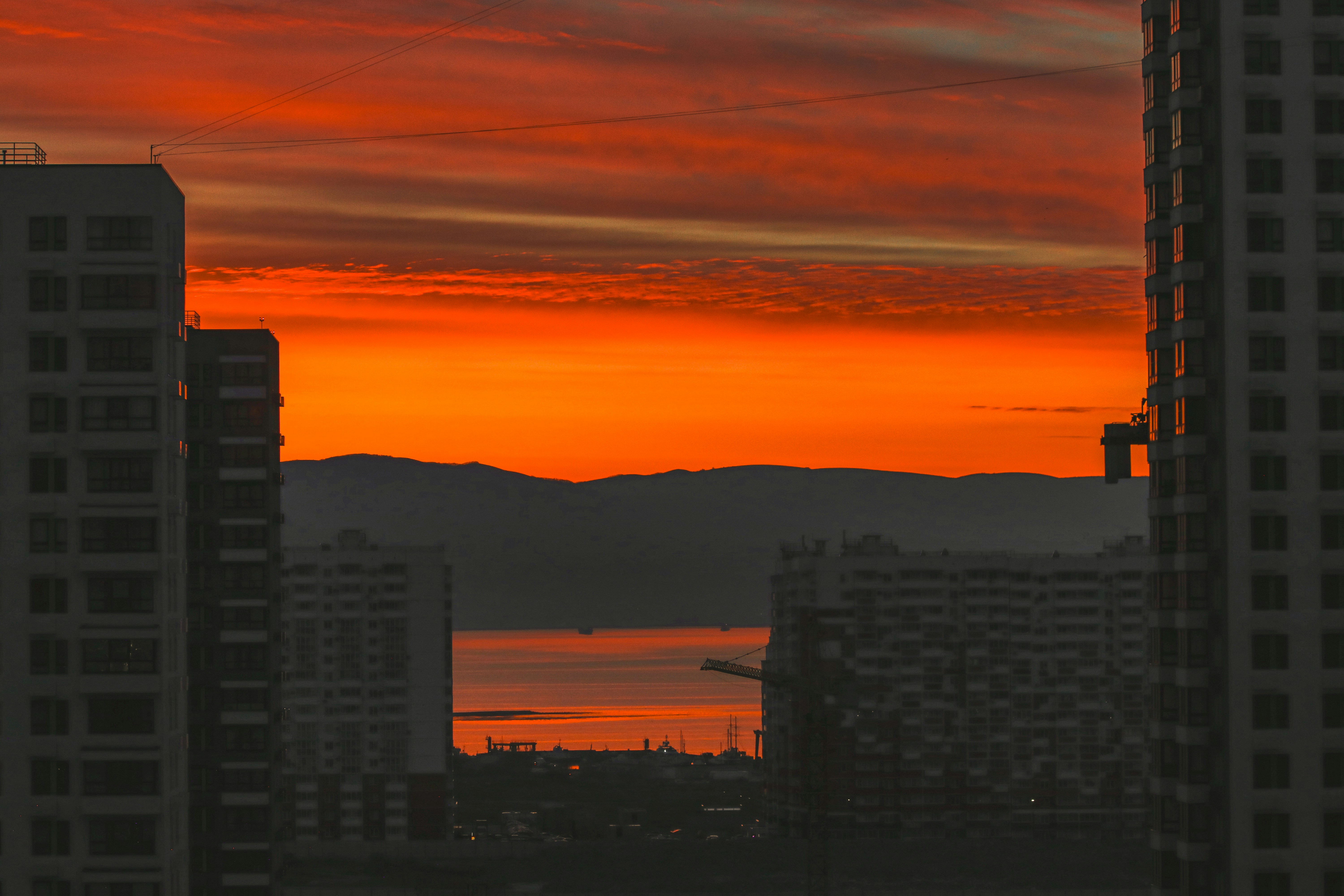 Silhouetted city buildings frame a vivid orange sunset over distant hills.