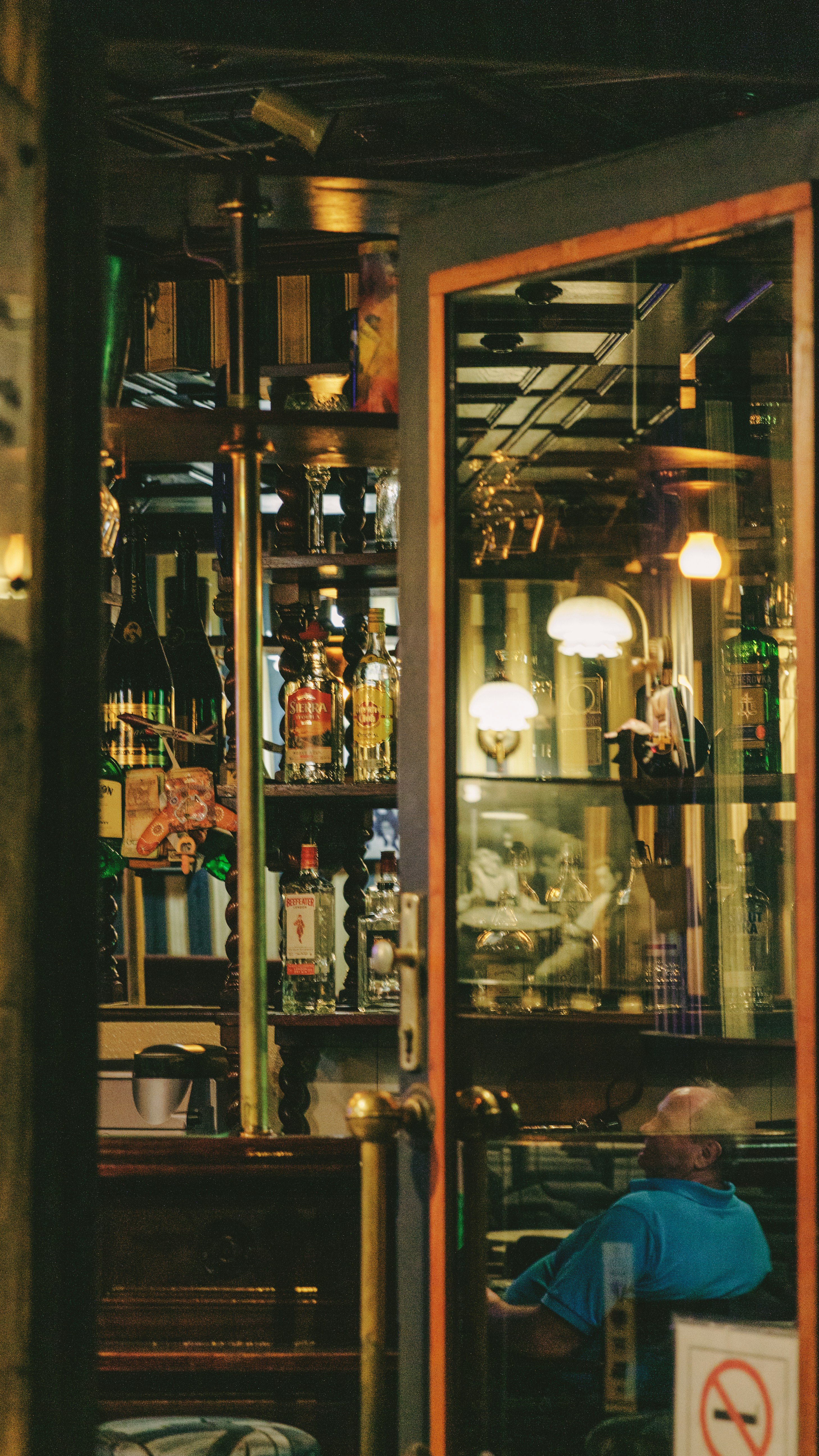 A cozy bar scene captured through a glass door, showcasing shelves filled with various bottles and a solitary patron in a blue shirt. 