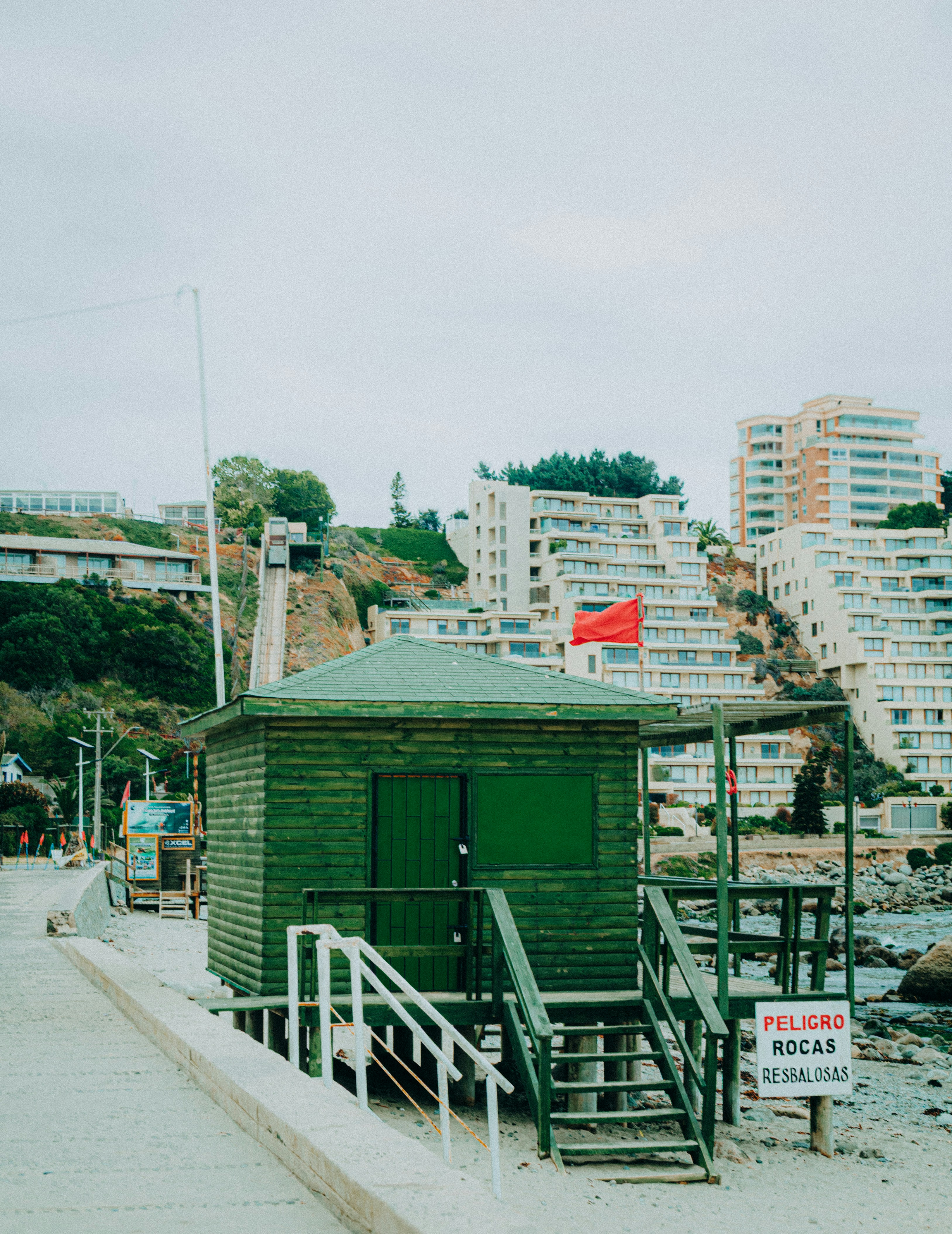 Green lifeguard station with red flag on a seaside walkway, cityscape and hills in the background.