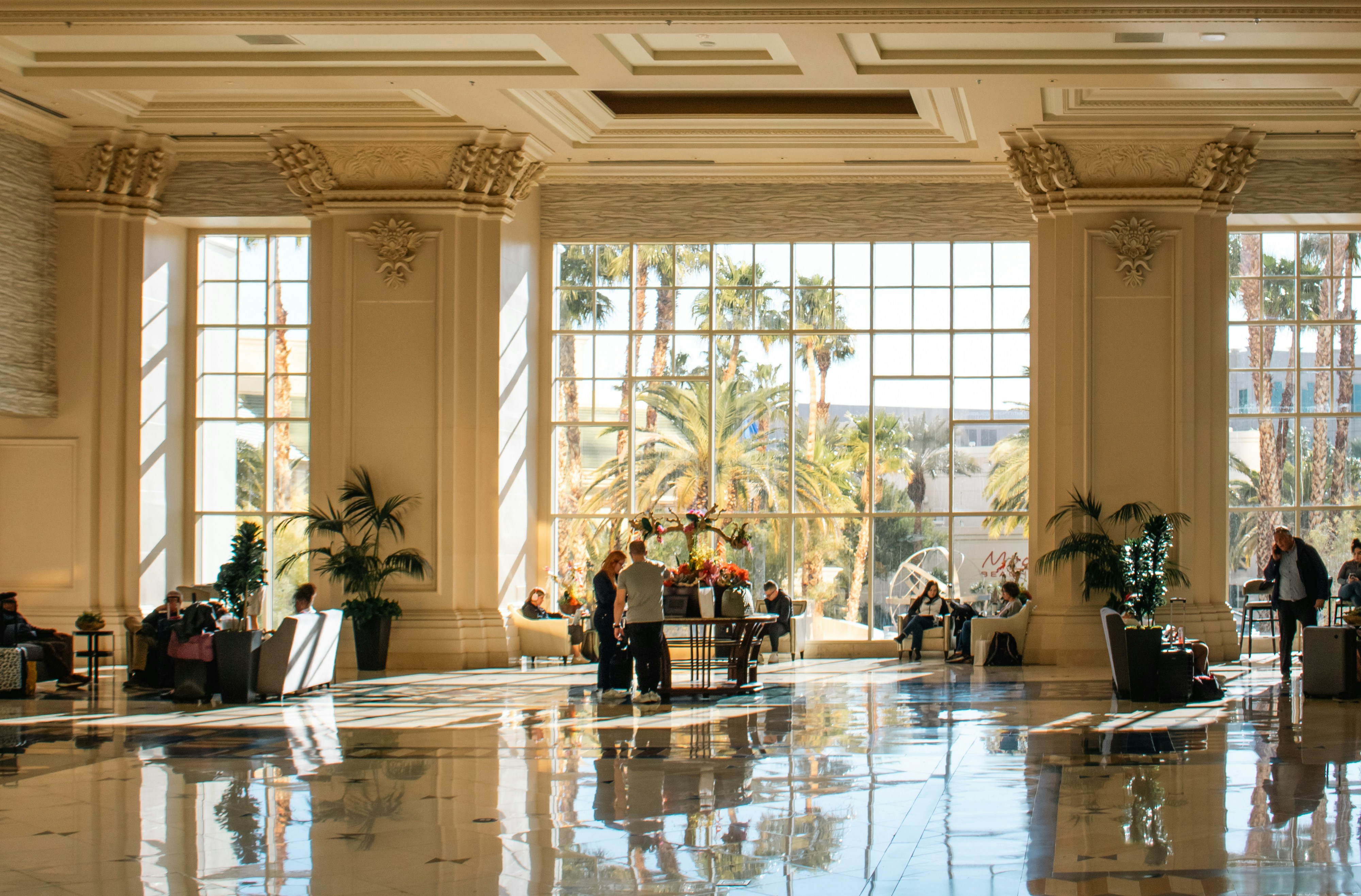 Sunlight streams through large windows, illuminating polished floors and ornate columns in a hotel lobby.