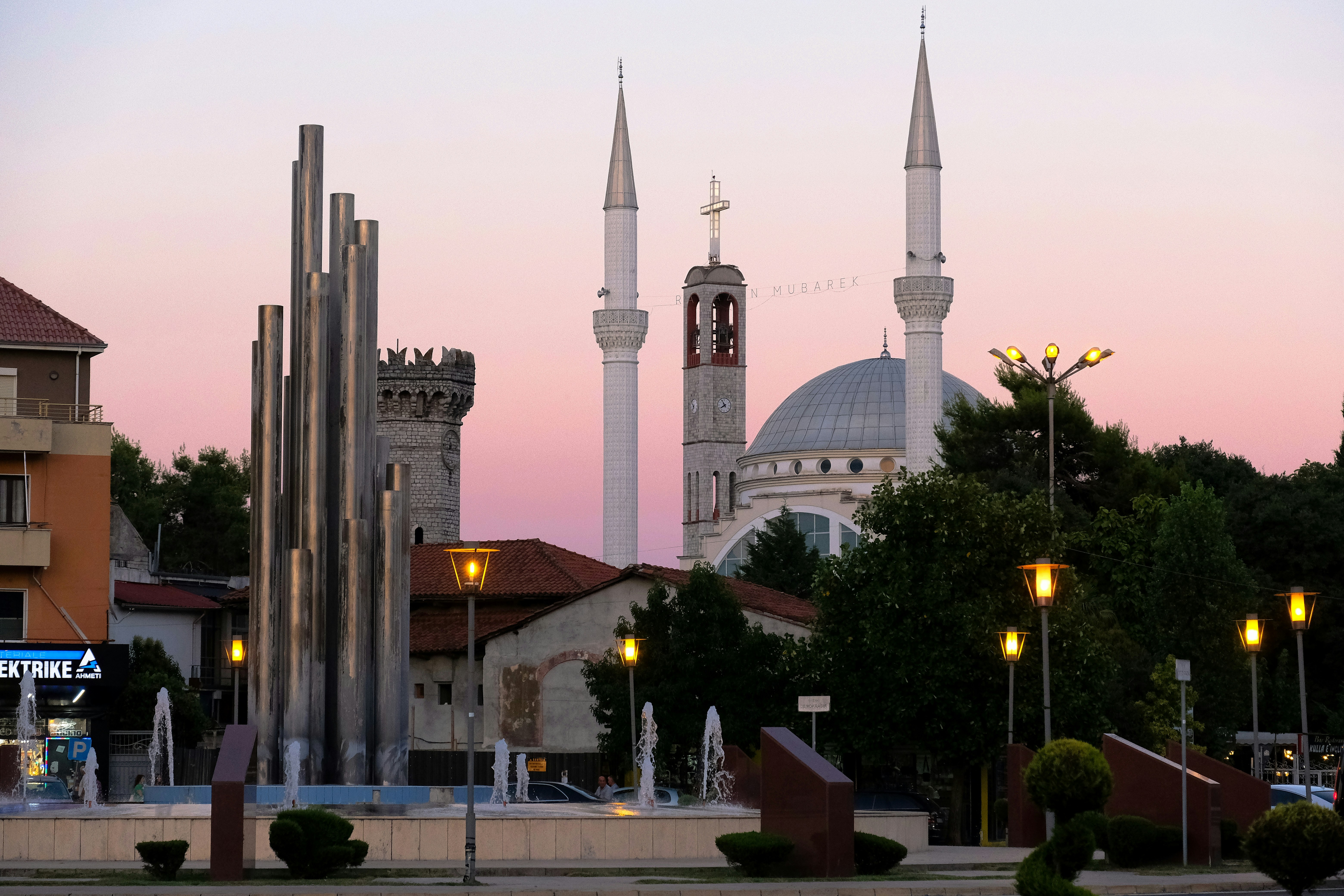 Mosque with two minarets, historic stone tower, and modern monument under a pink evening sky.