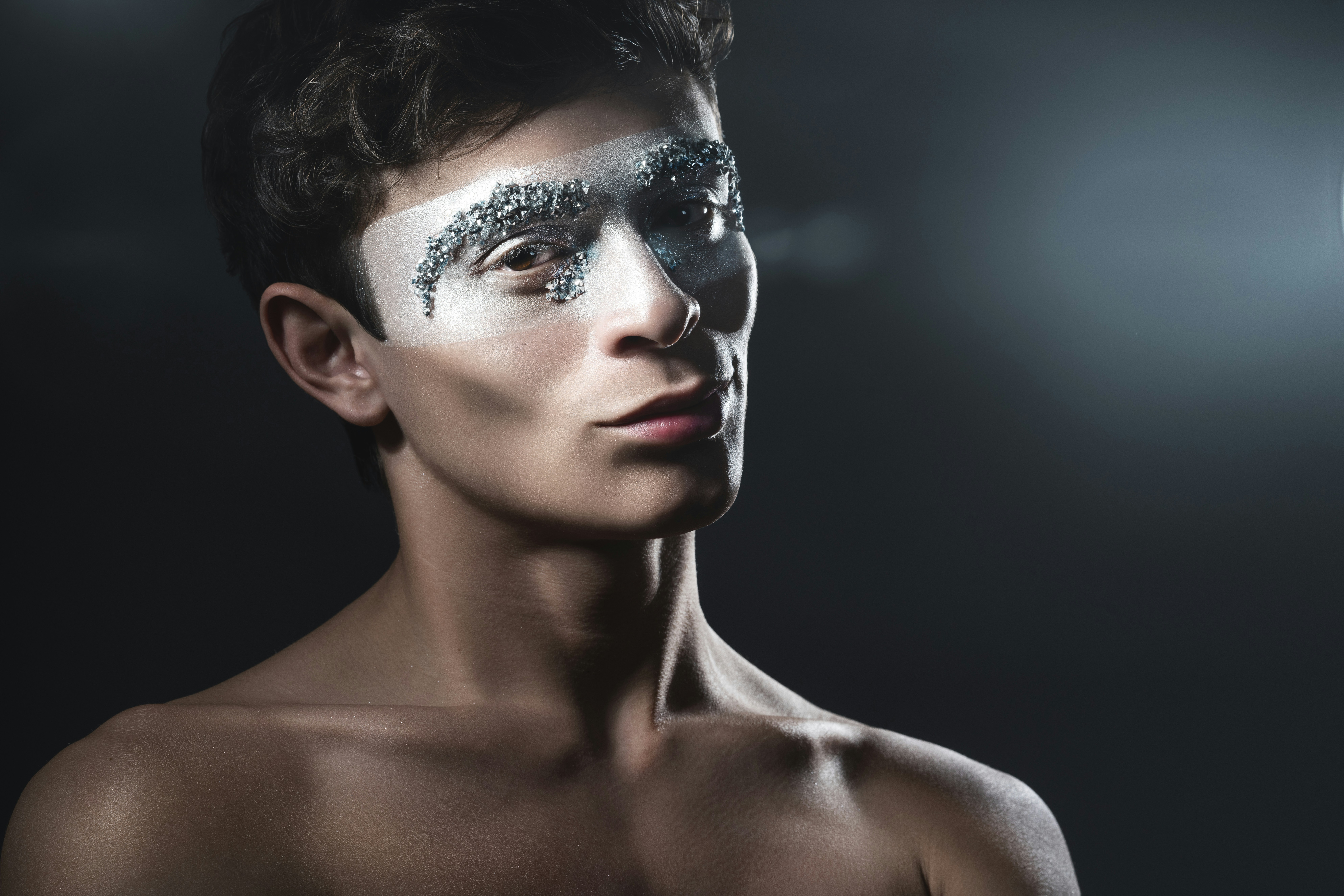Person with silver and black face paint gazes into the distance under dramatic lighting.