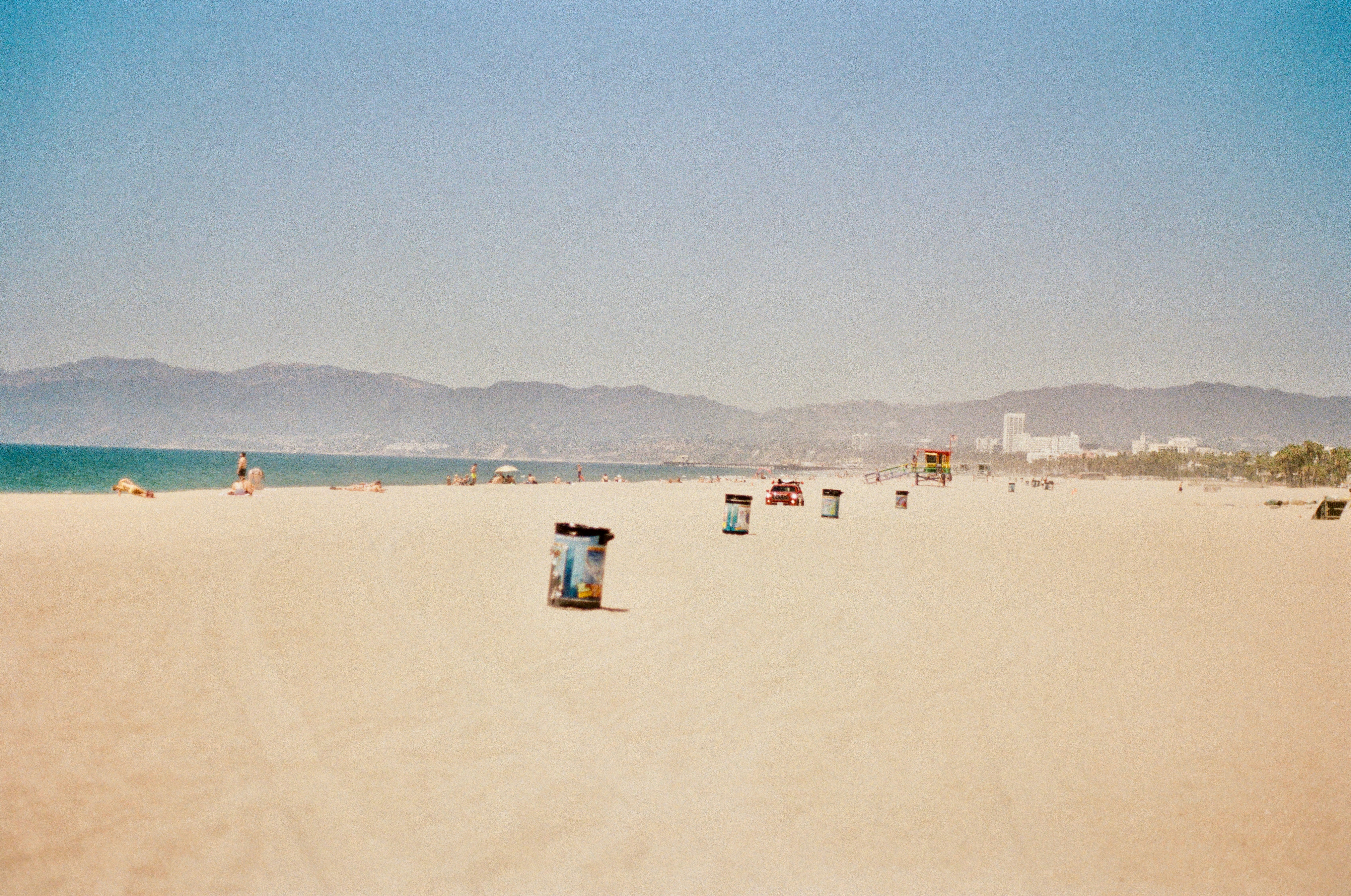 Wide sandy beach with scattered trash bins under a clear blue sky.