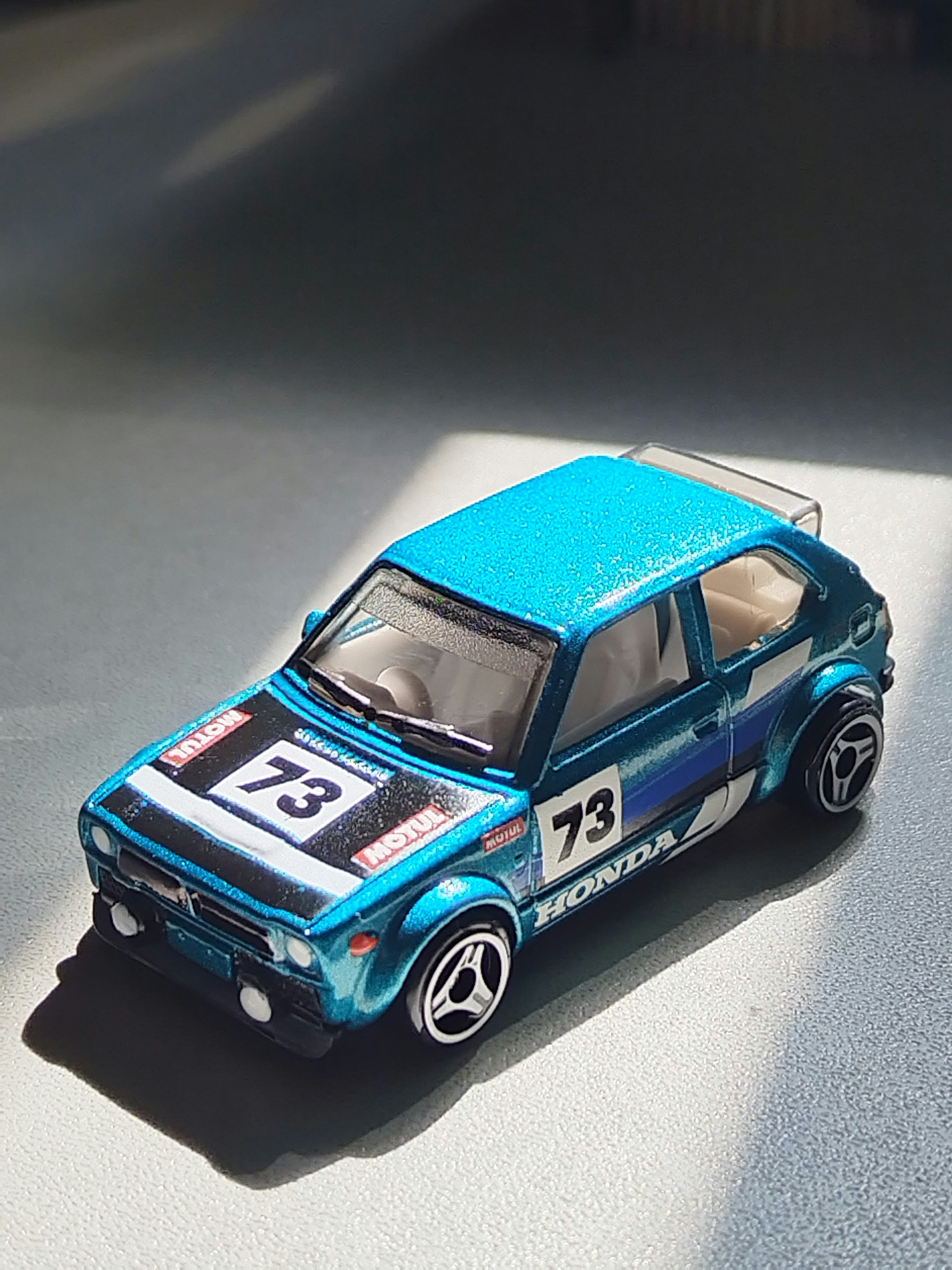 Close-up photograph of a blue die-cast Honda rally car toy with white racing numbers on a sunlit tabletop.