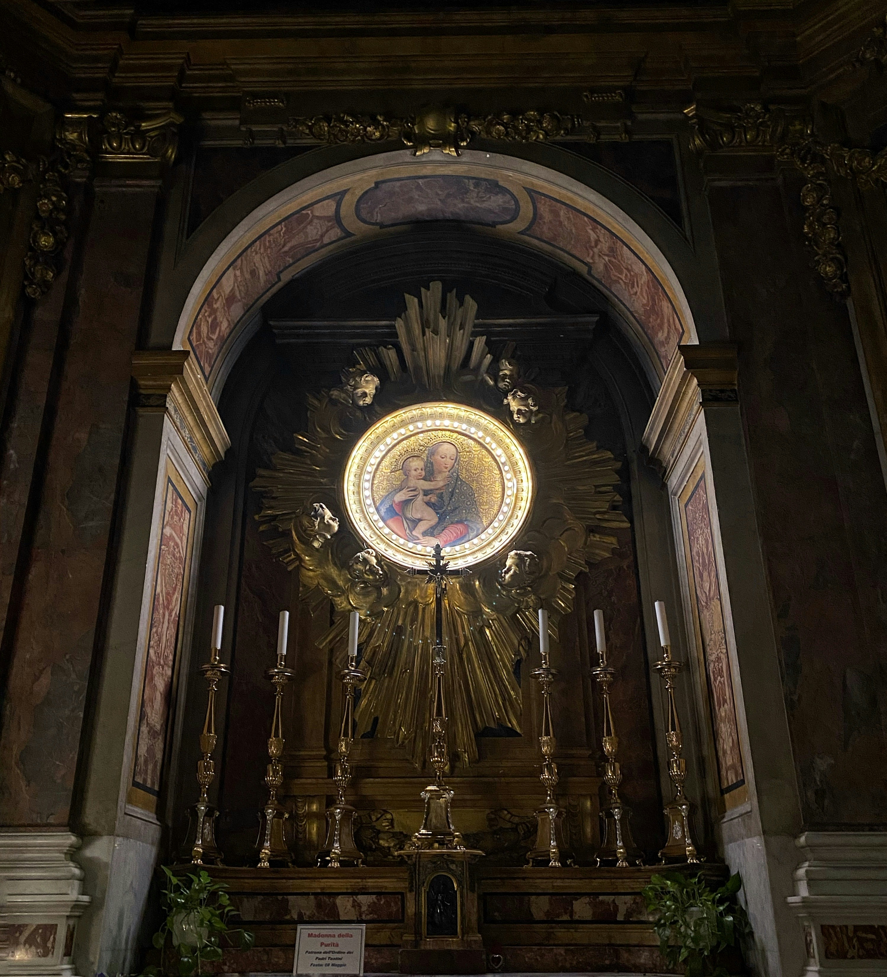 Ornate religious altar with golden candlesticks and a radiant circular artwork in a dimly lit chapel.