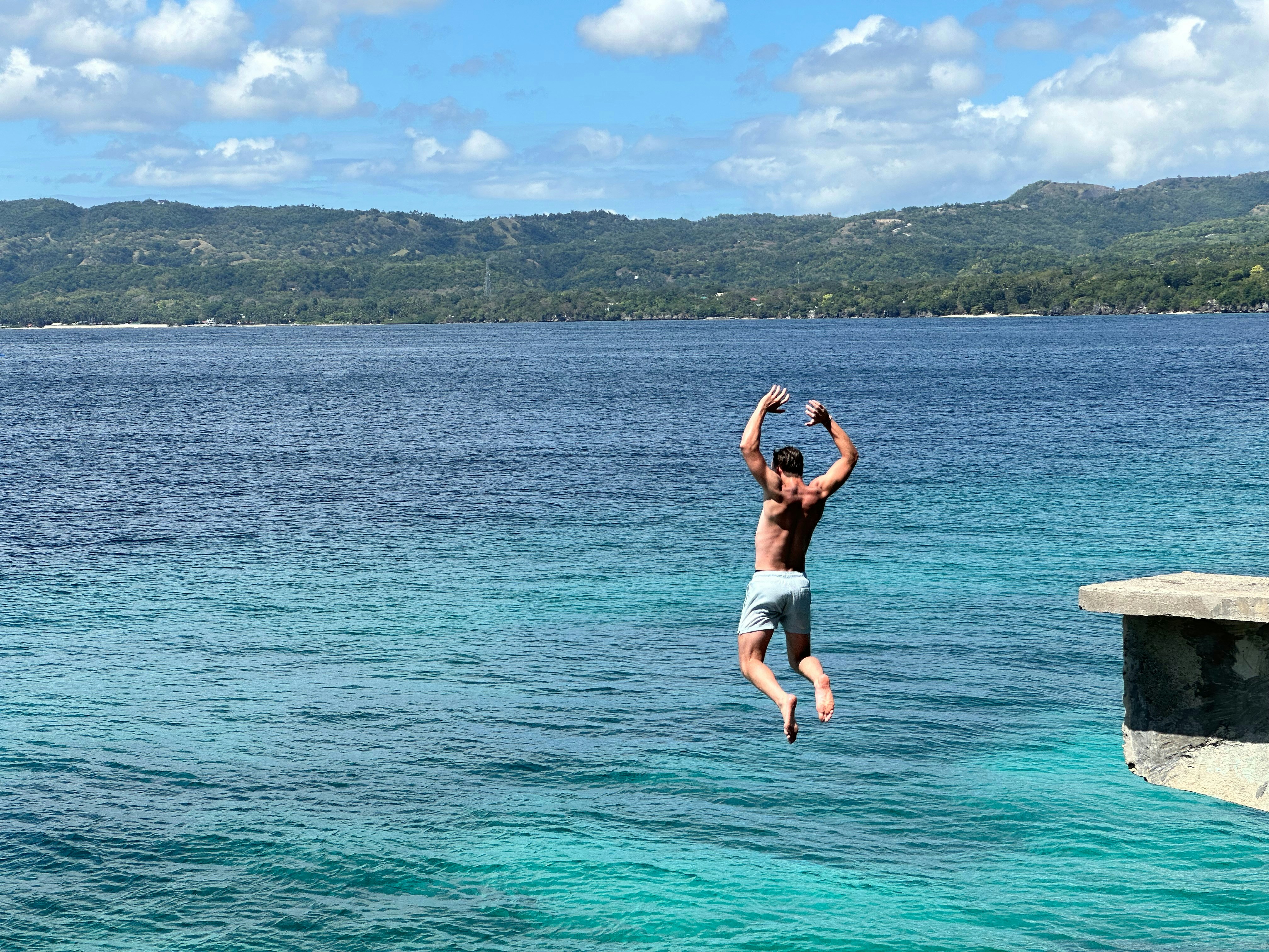 Man mid-jump from a cliff into vibrant blue ocean under a clear sky.
