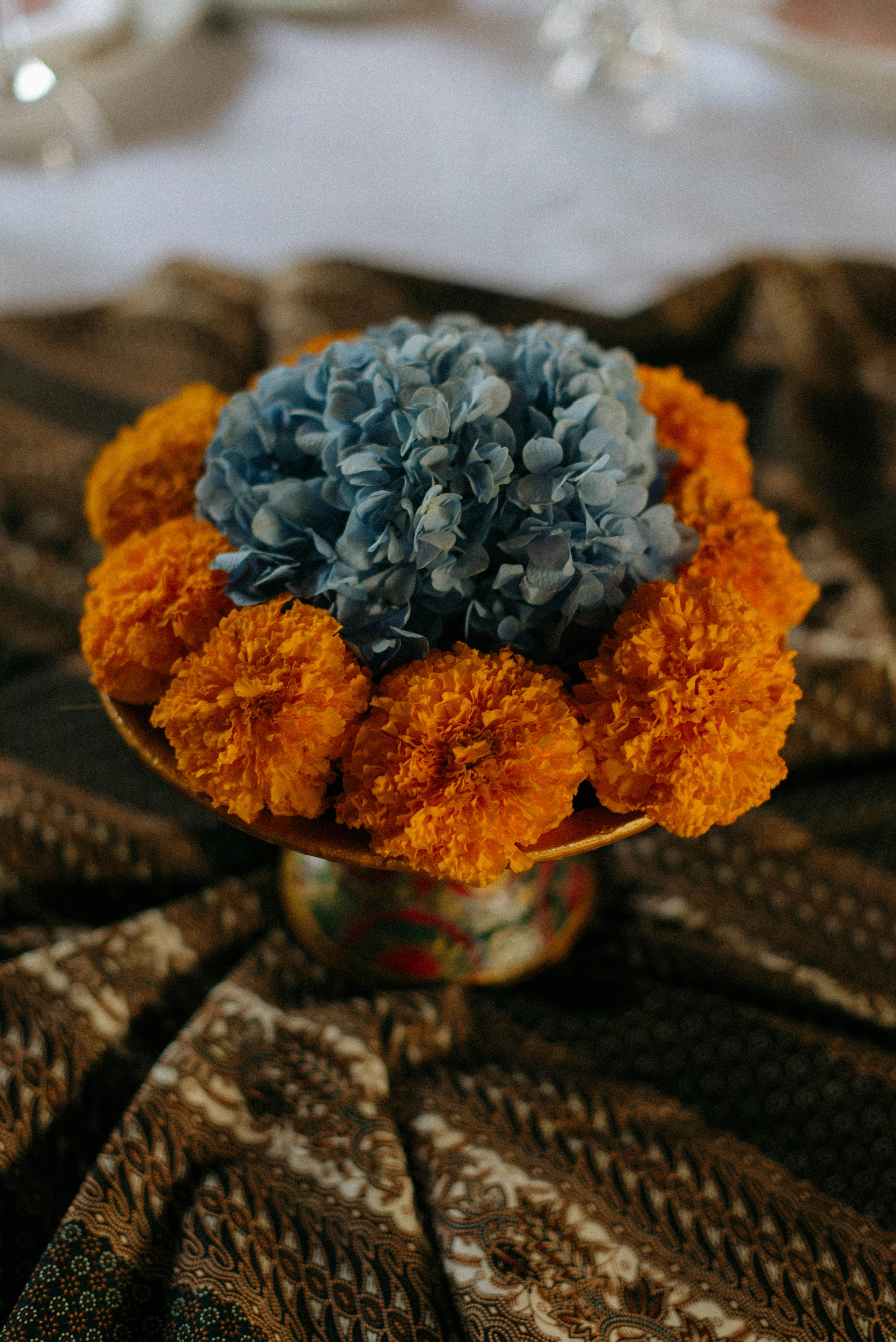Blue hydrangea surrounded by orange marigolds in a decorative bowl on patterned fabric.