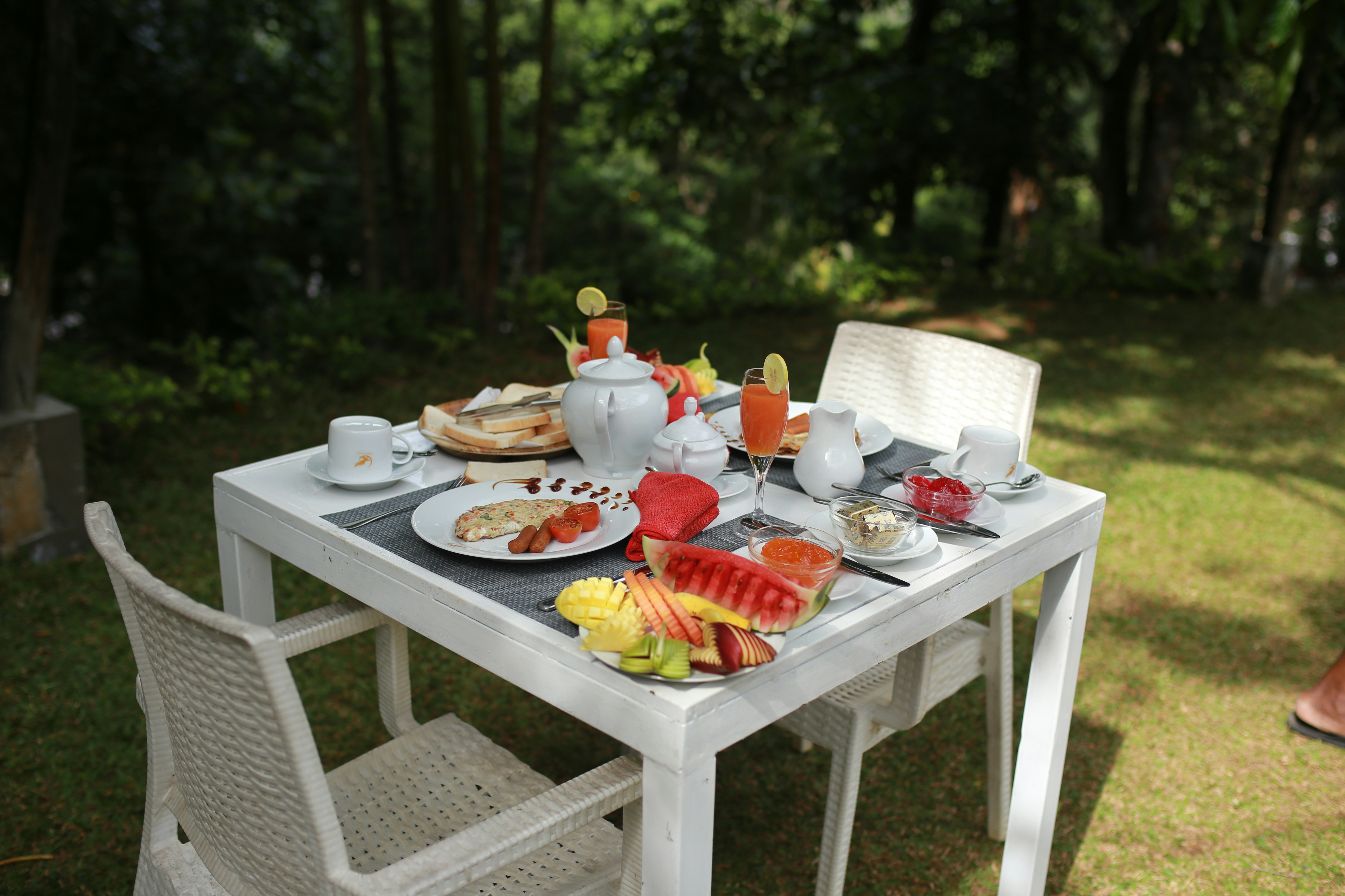 Outdoor dining table set with a variety of colorful fruits and breakfast dishes under dappled sunlight.