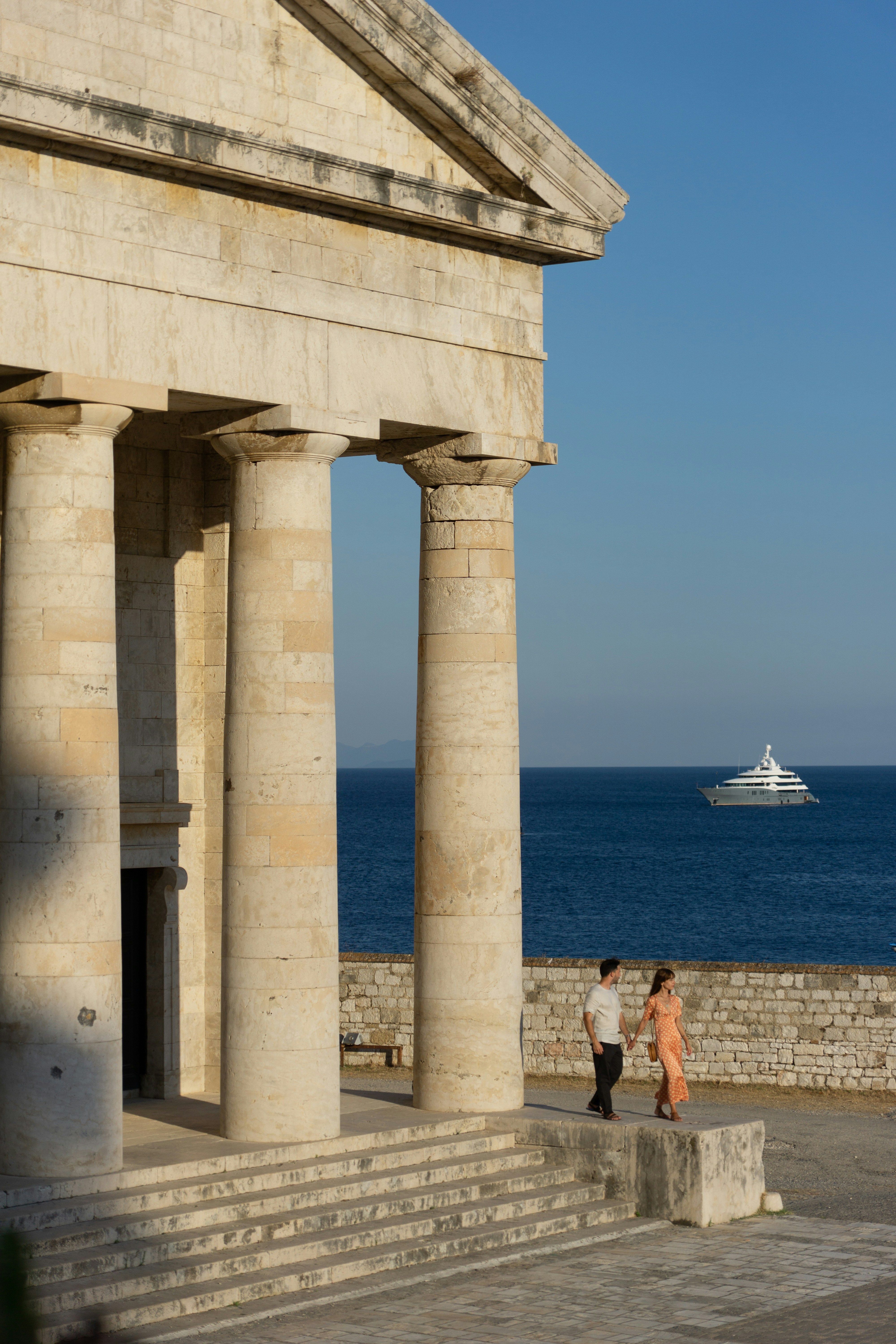 Couple standing near ancient columns with a yacht on the horizon under a clear blue sky.
