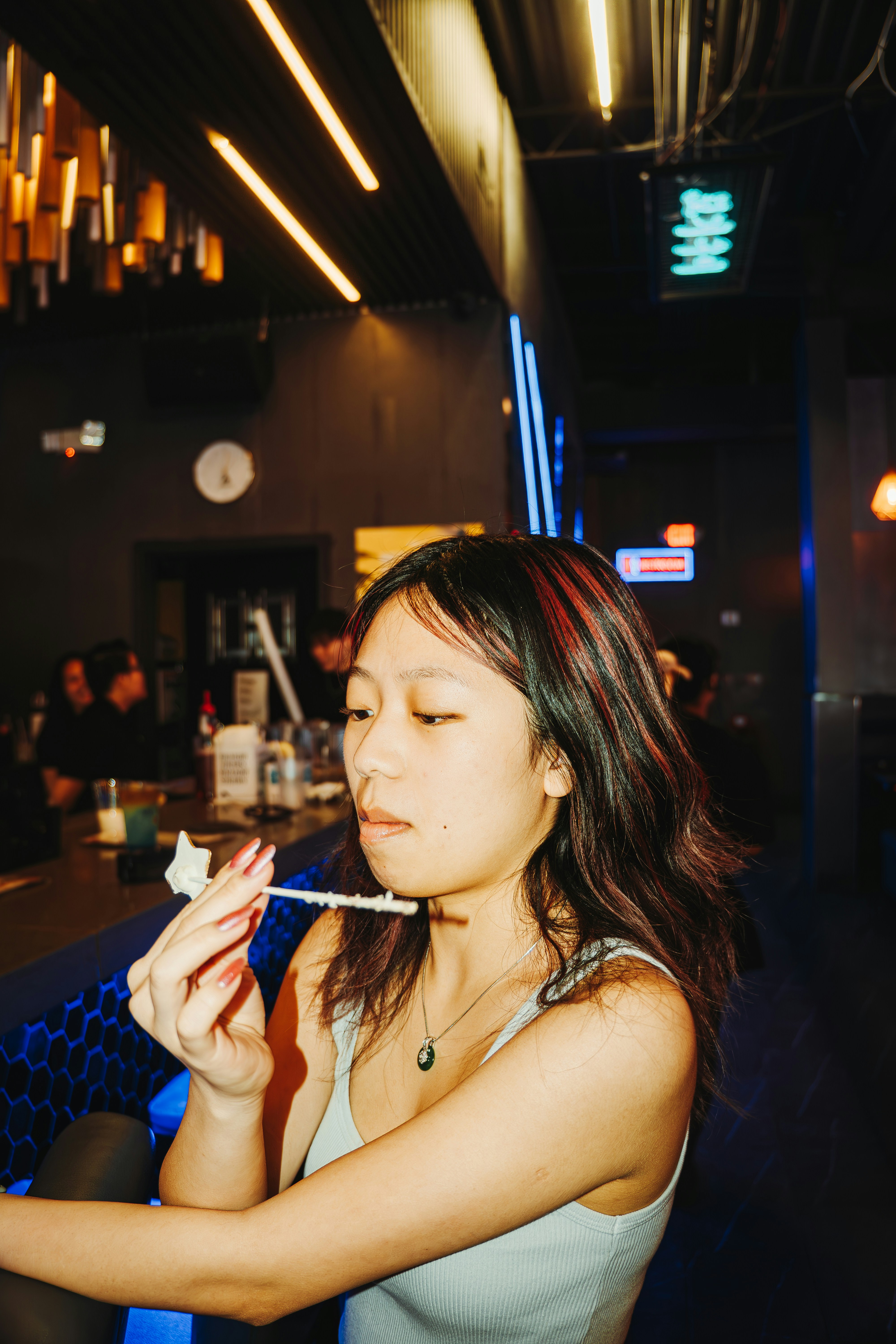 Young woman savoring a dish at a vibrant bar, illuminated by colorful lights and modern decor.