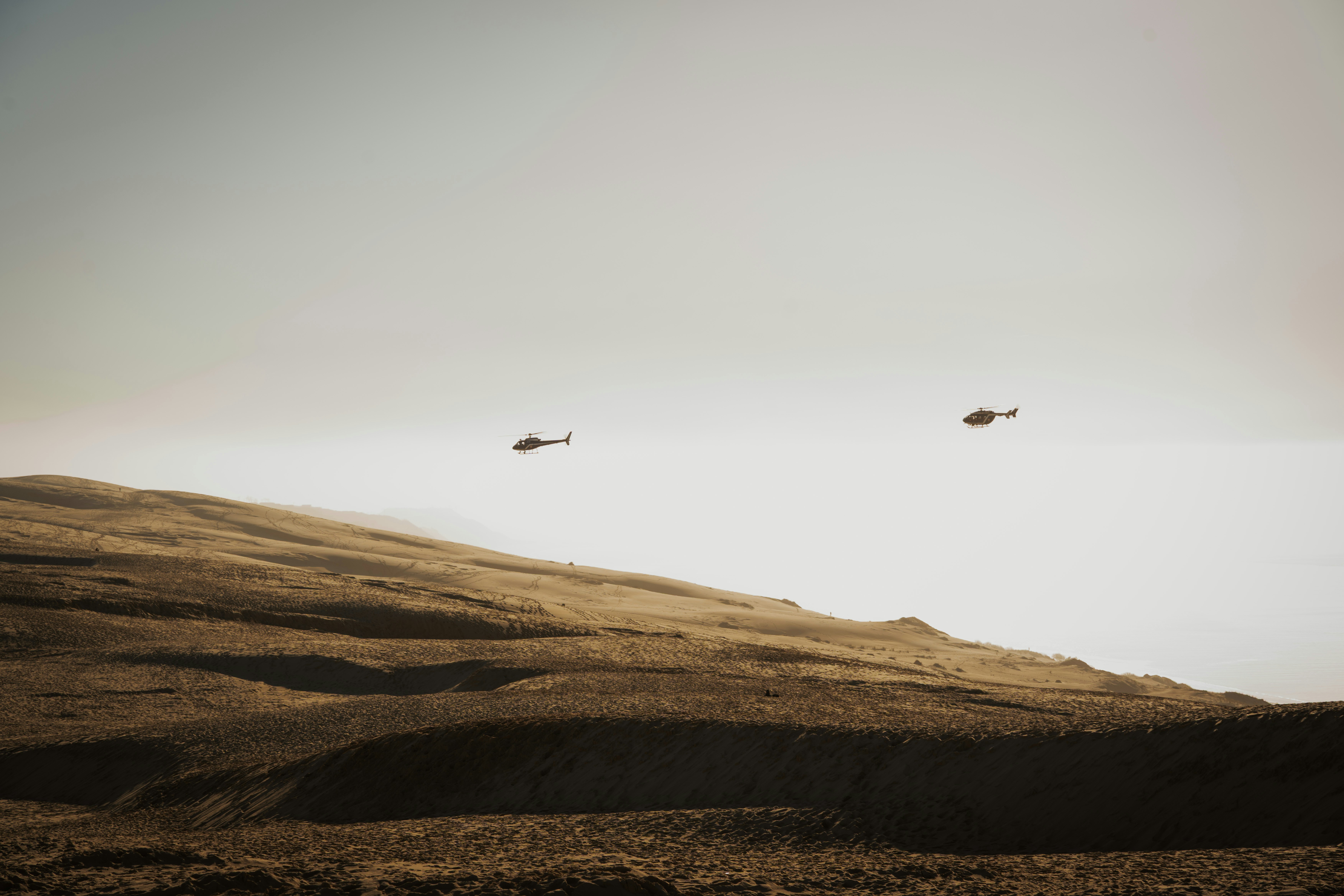 Two helicopters flying over expansive sand dunes under a hazy sky.