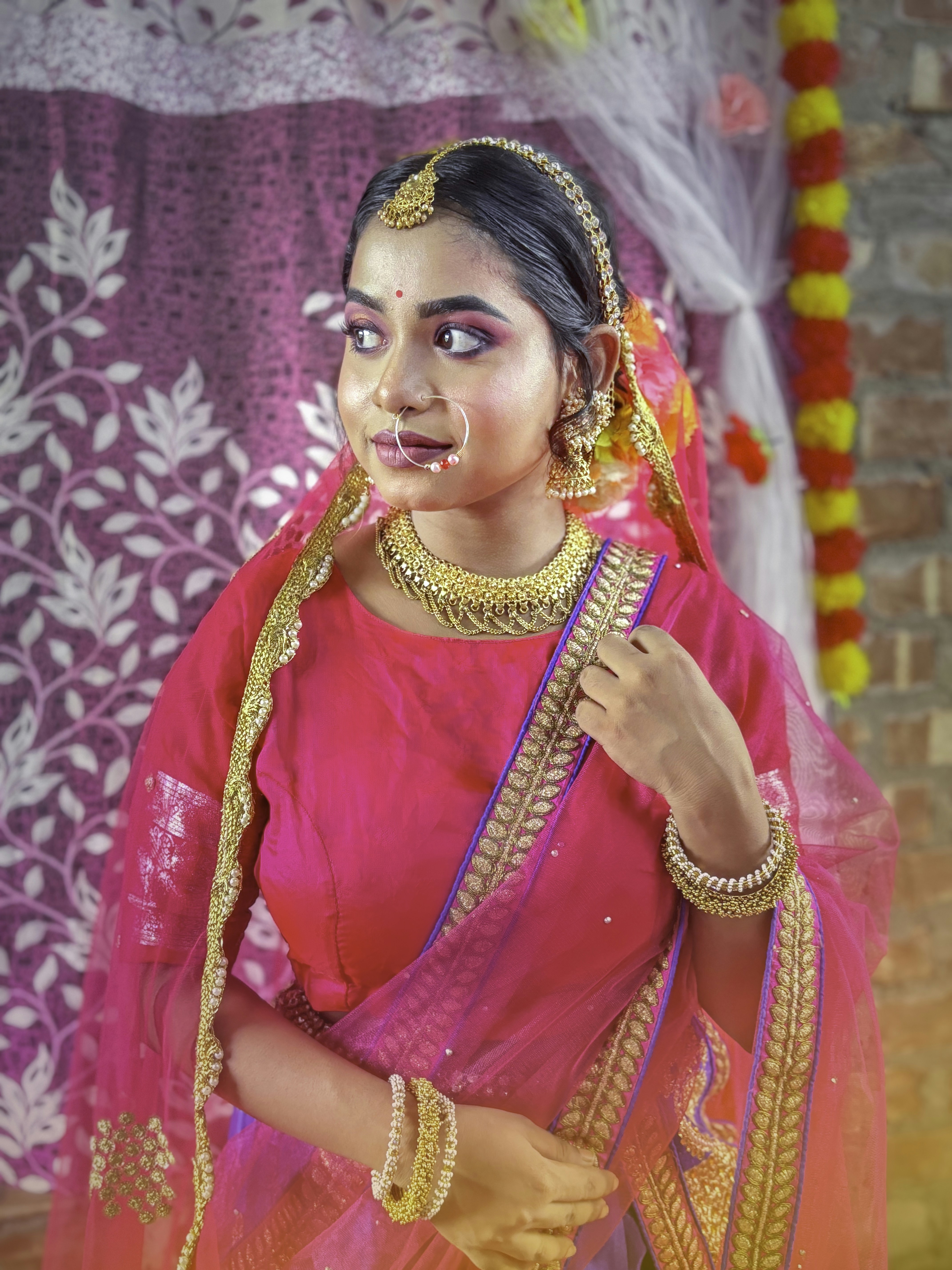 Bride in vibrant pink and gold traditional attire, adorned with intricate jewelry against a decorative backdrop.