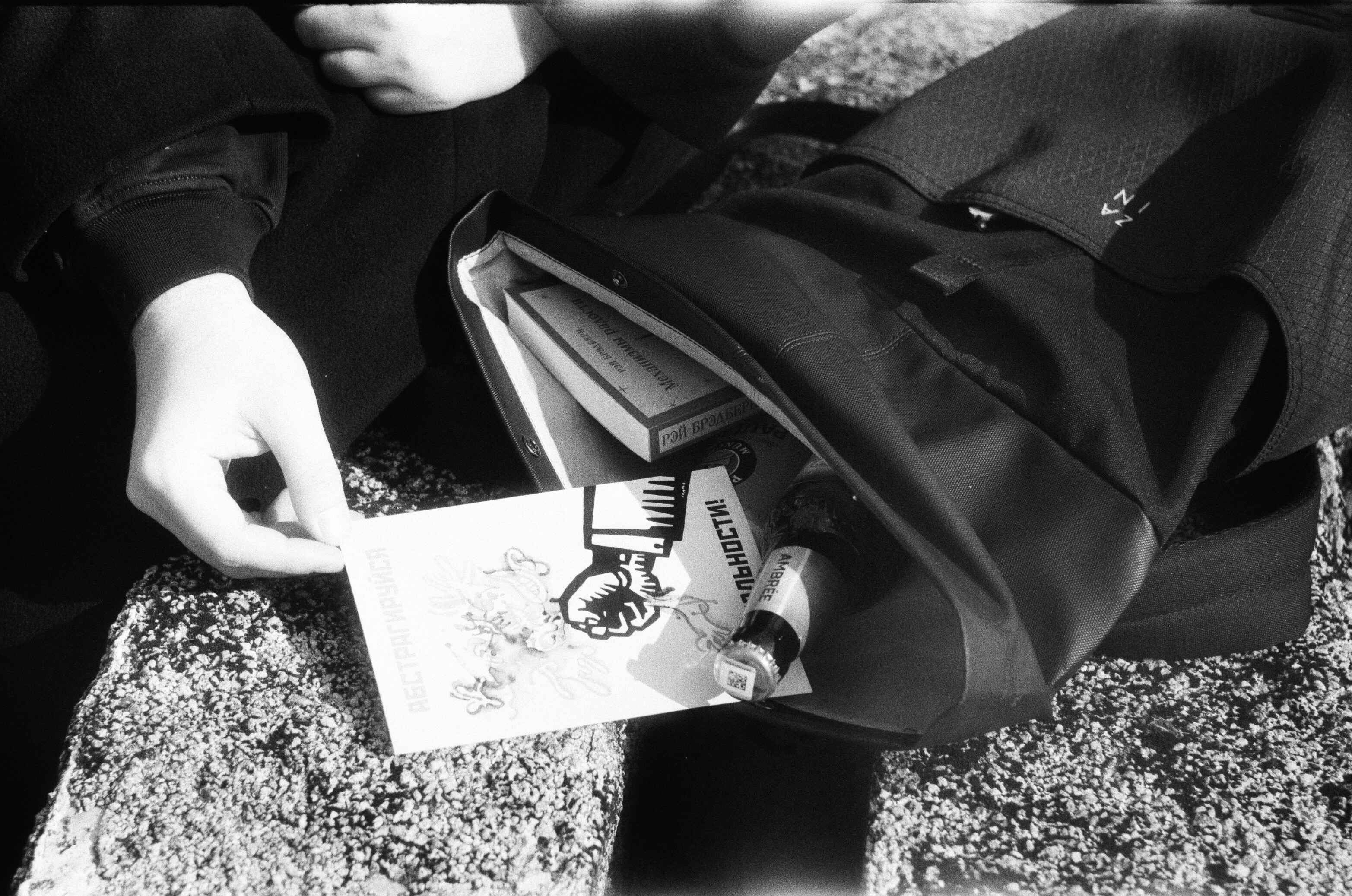 Black and white photo of a person holding a postcard next to an open backpack with books and a camera inside.